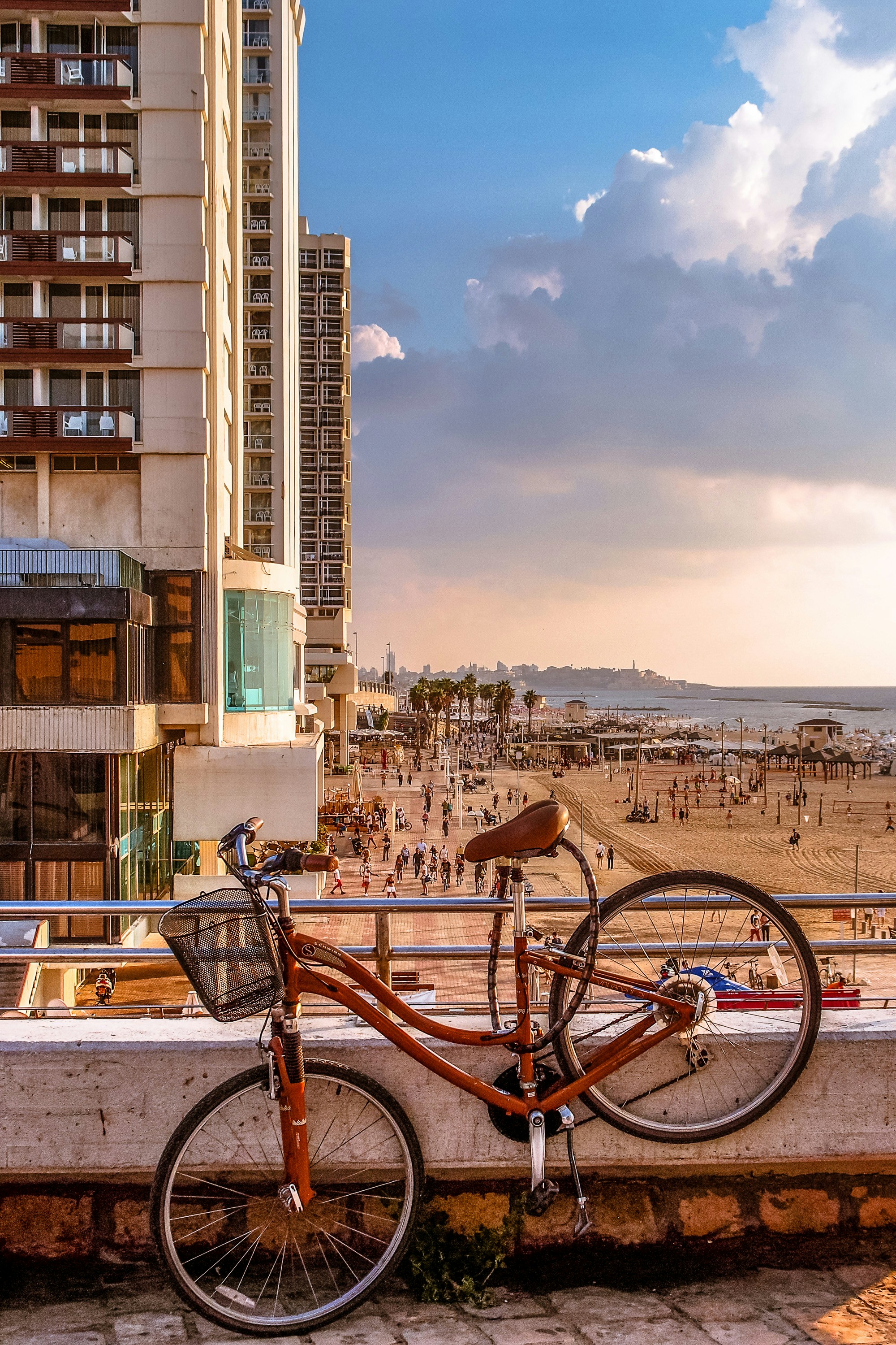 Orange bicycle parked on a railing overlooks a bustling beach. Tall apartment blocks line the shore as the sun sets over the calm sea.