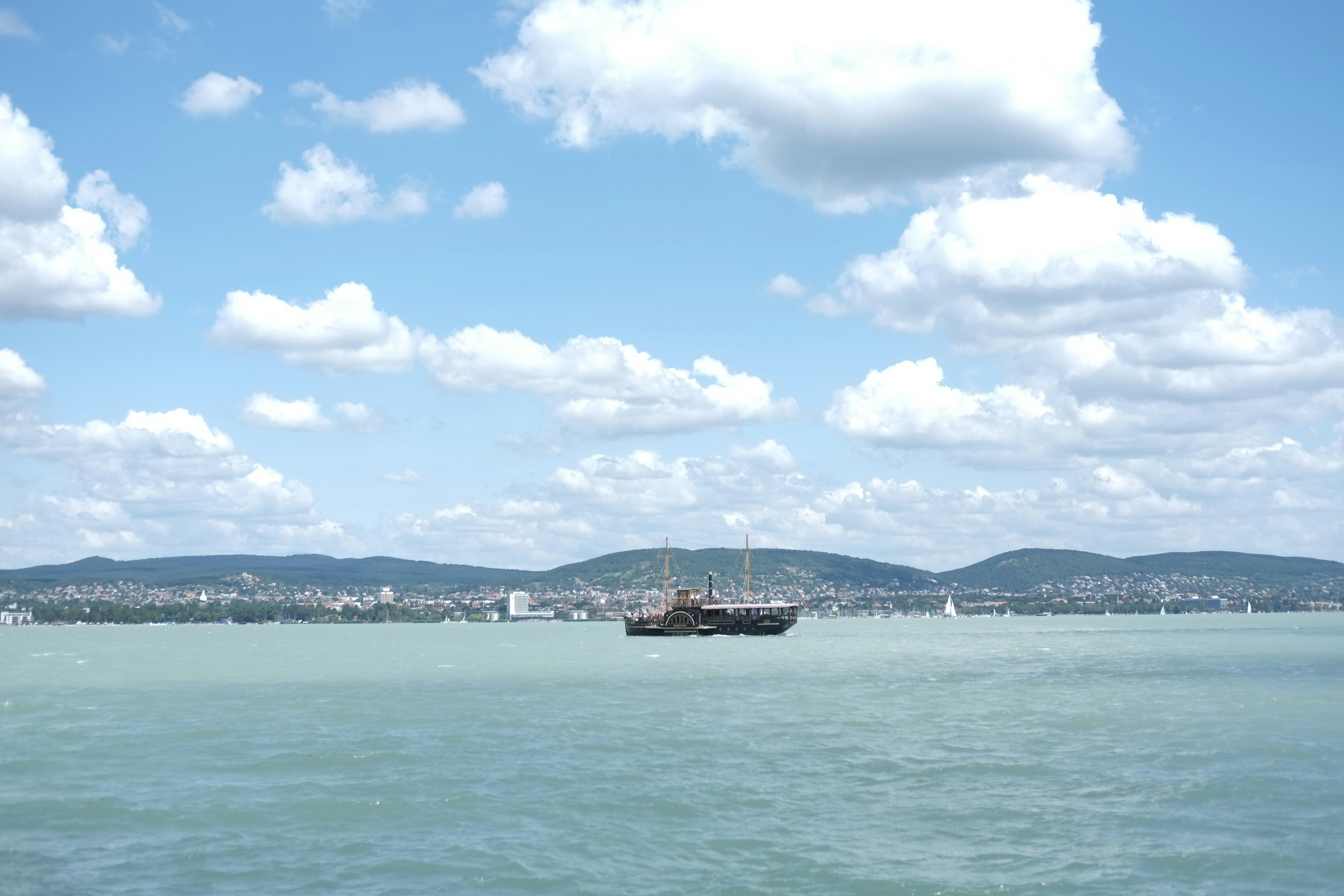 A vintage sailing ship glides across a tranquil lake under a sky filled with fluffy clouds, framed by distant hills. The scene evokes a sense of adventure and peace.