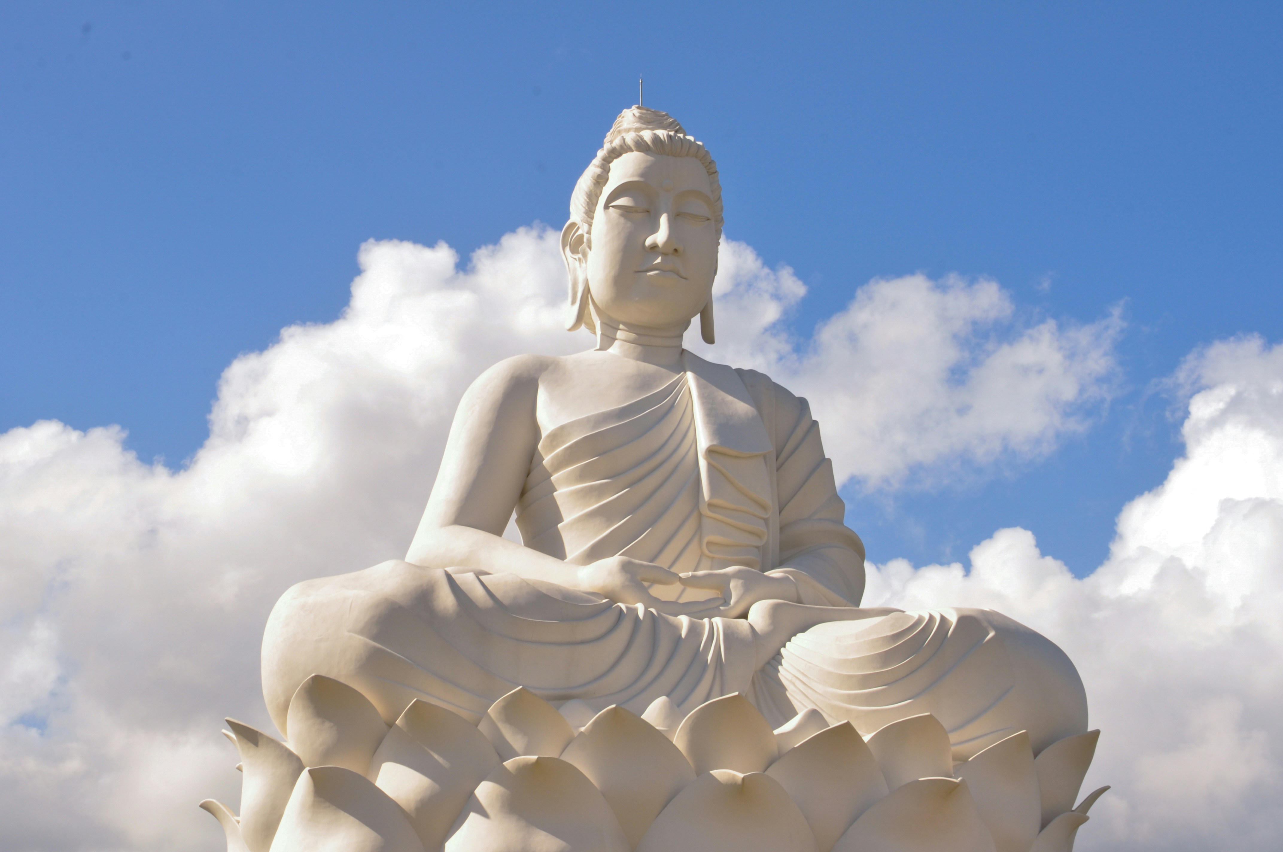 Large white Buddha statue seated against a backdrop of fluffy clouds and clear blue sky.