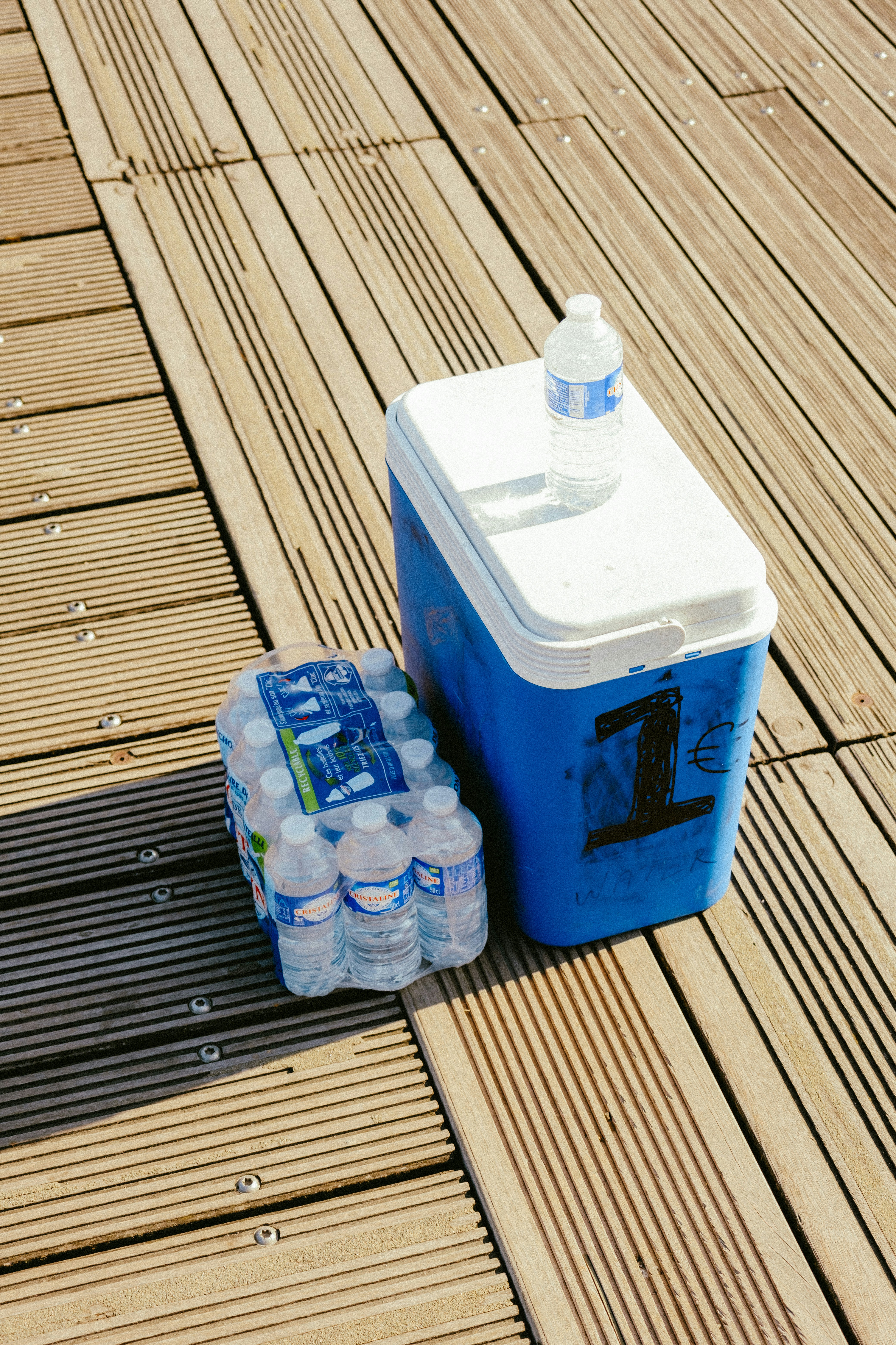 A blue cooler with a water bottle on top sits beside a pack of bottled water on a wooden deck.