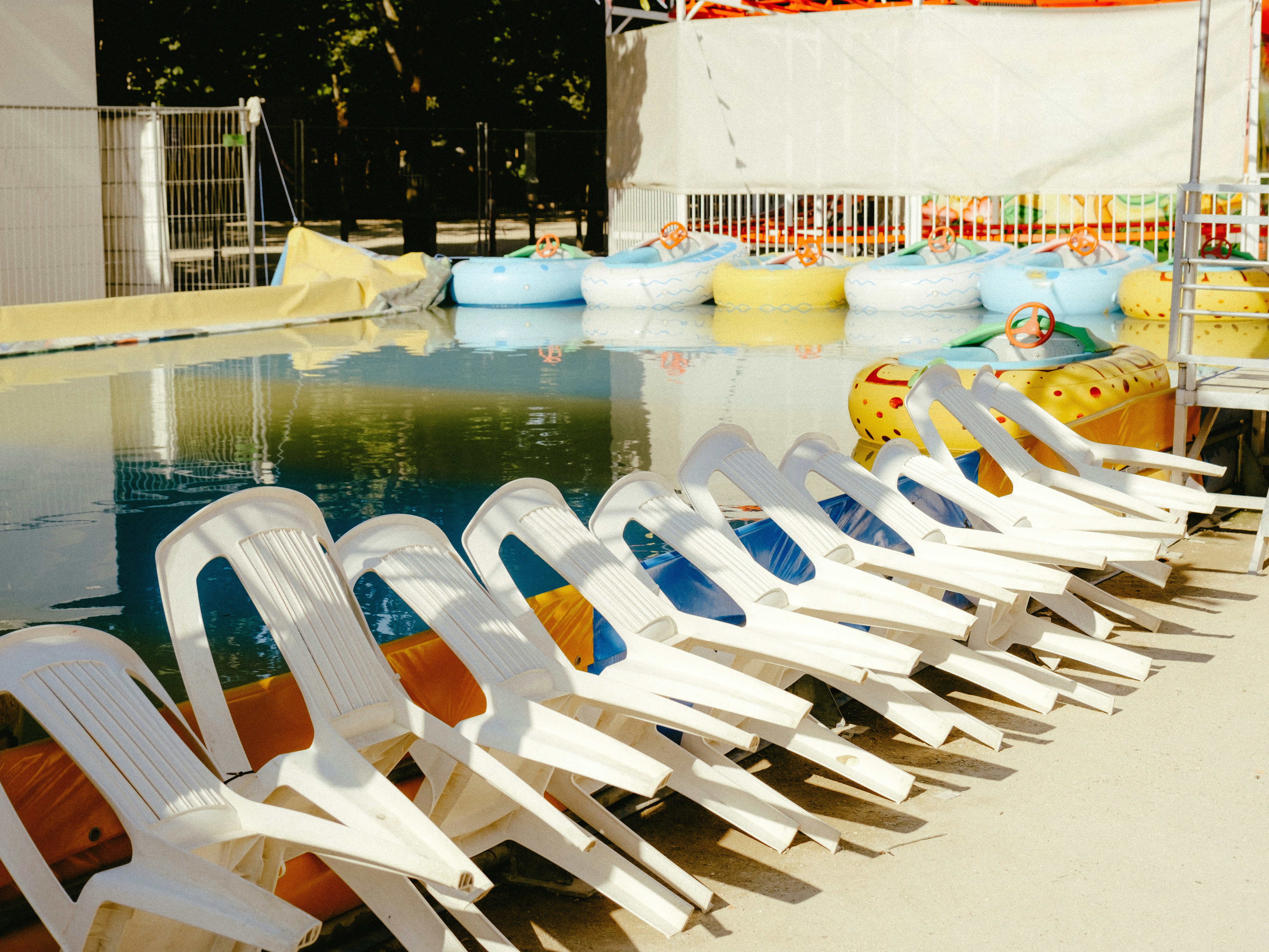 White plastic chairs line the edge of a tranquil pool, surrounded by colorful inflatable floats under a bright sunlit sky.