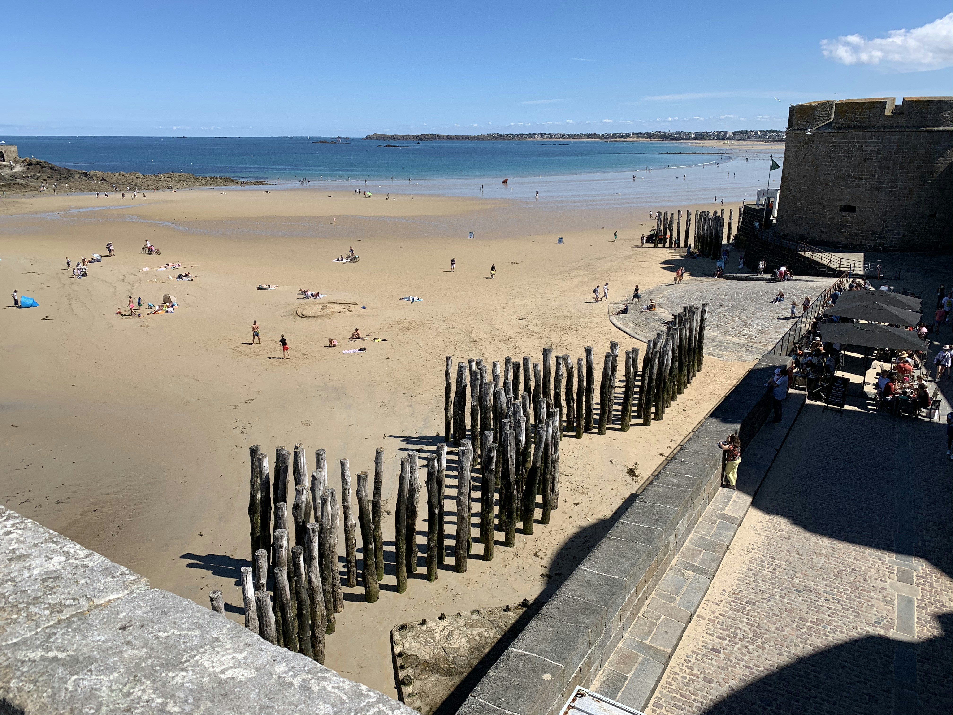 Sandy beach with scattered beachgoers, a historic stone wall, and wooden groynes under a clear blue sky.