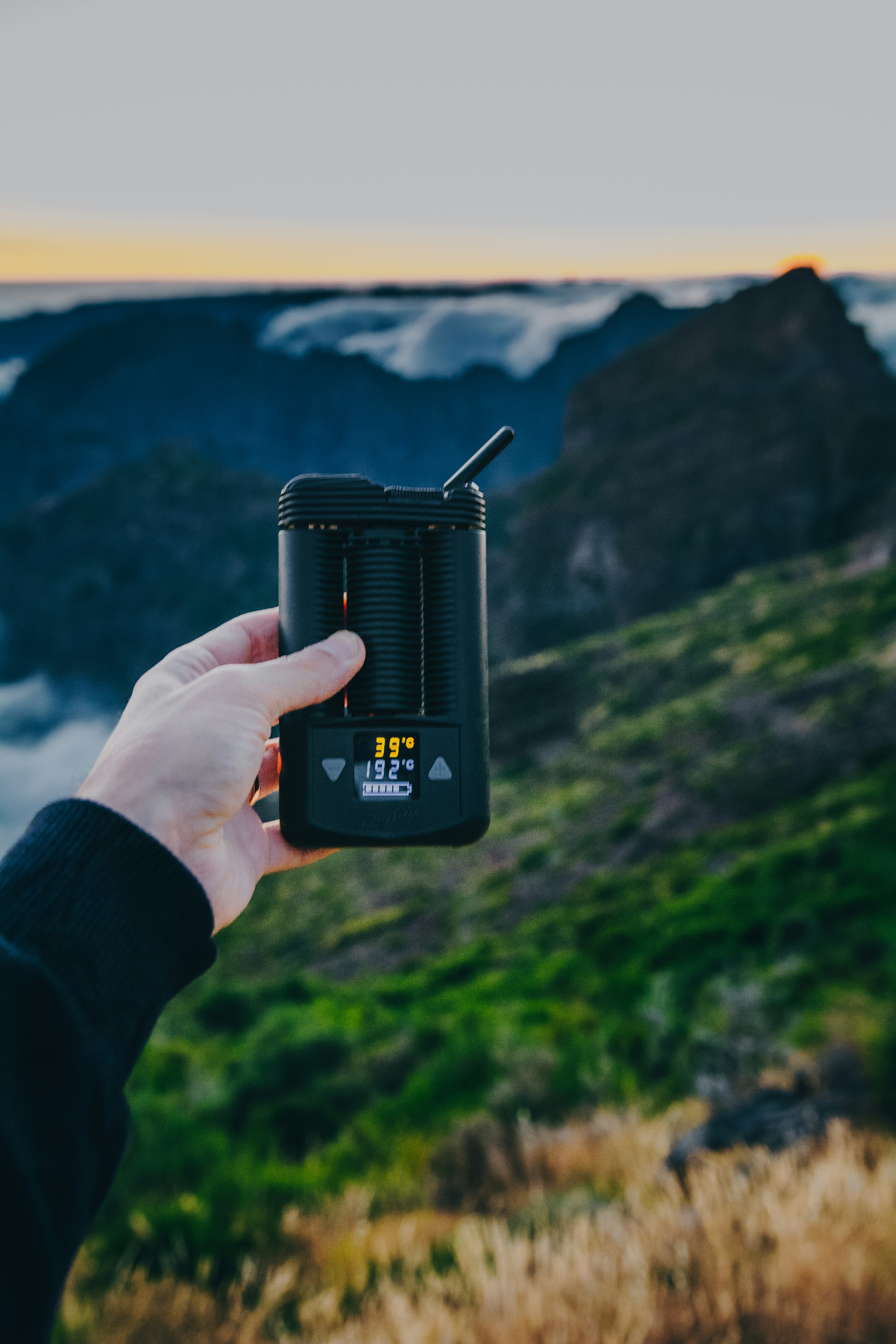 Hand holding a digital temperature gauge against a breathtaking mountainous backdrop at dusk.