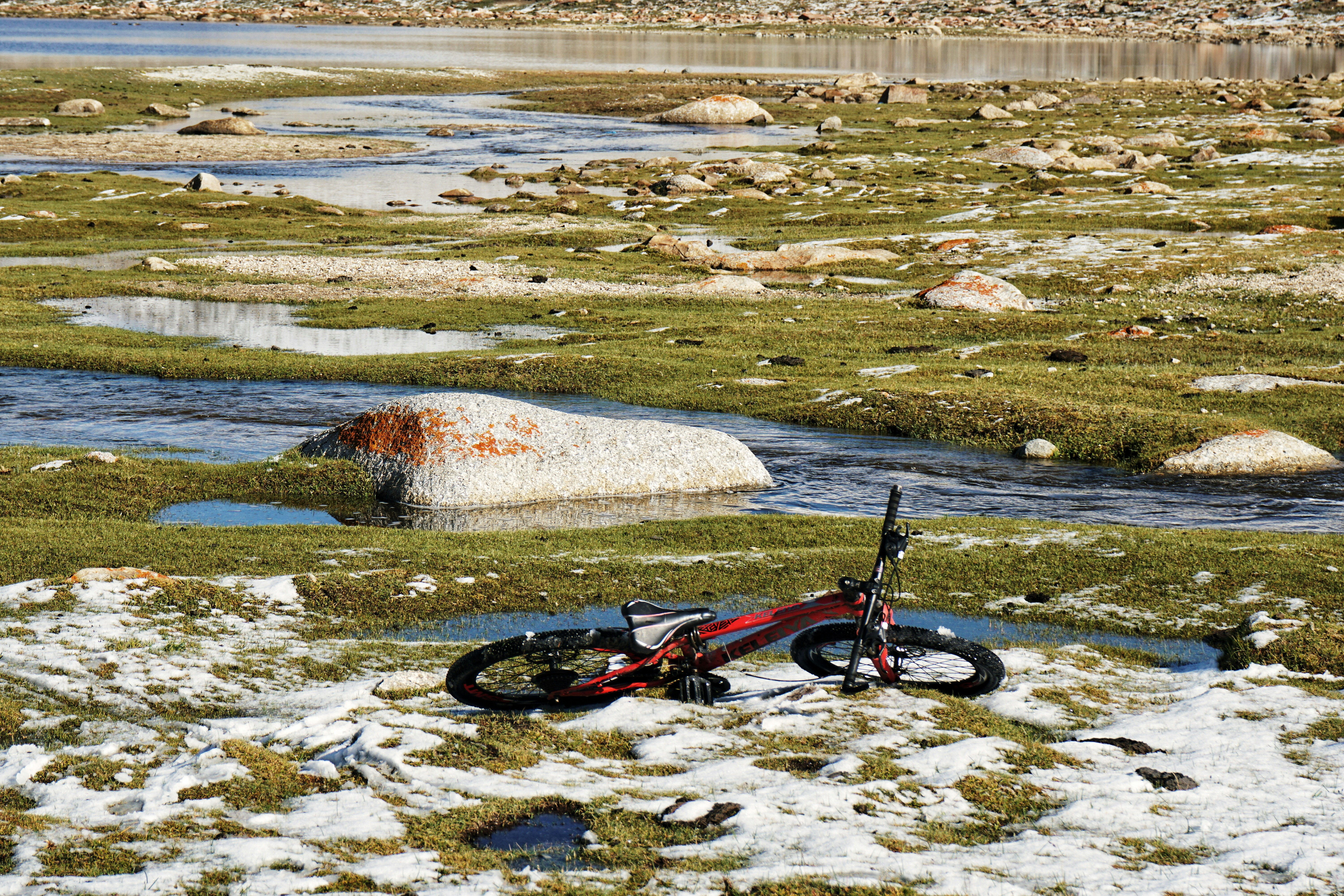 A red bicycle lies abandoned on a patch of snow and grass beside a stream, surrounded by large rocks and a serene landscape.