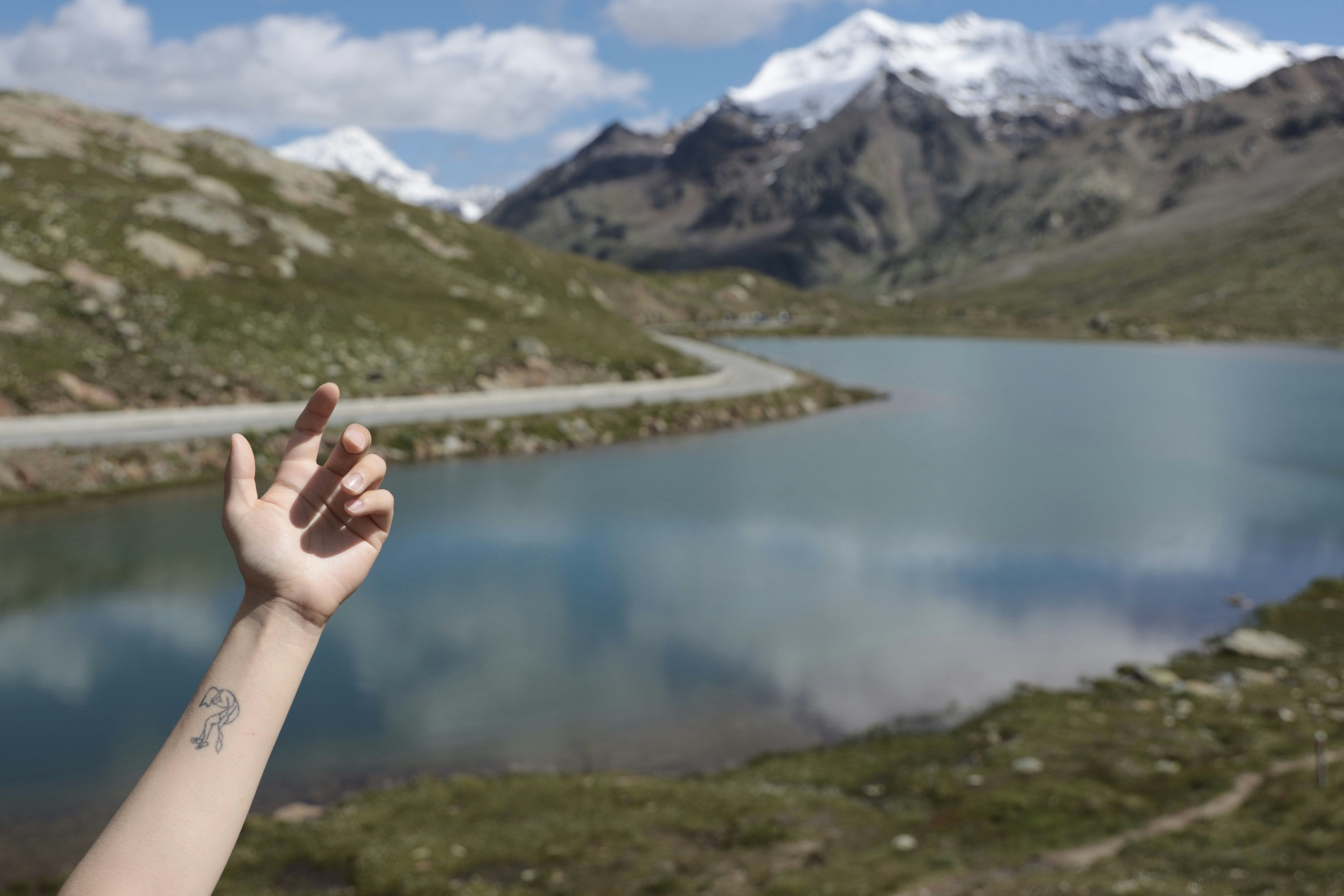 A hand reaching out towards a tranquil lake surrounded by majestic mountains, reflecting the sky's blue hues. The scene captures a moment of connection with nature.