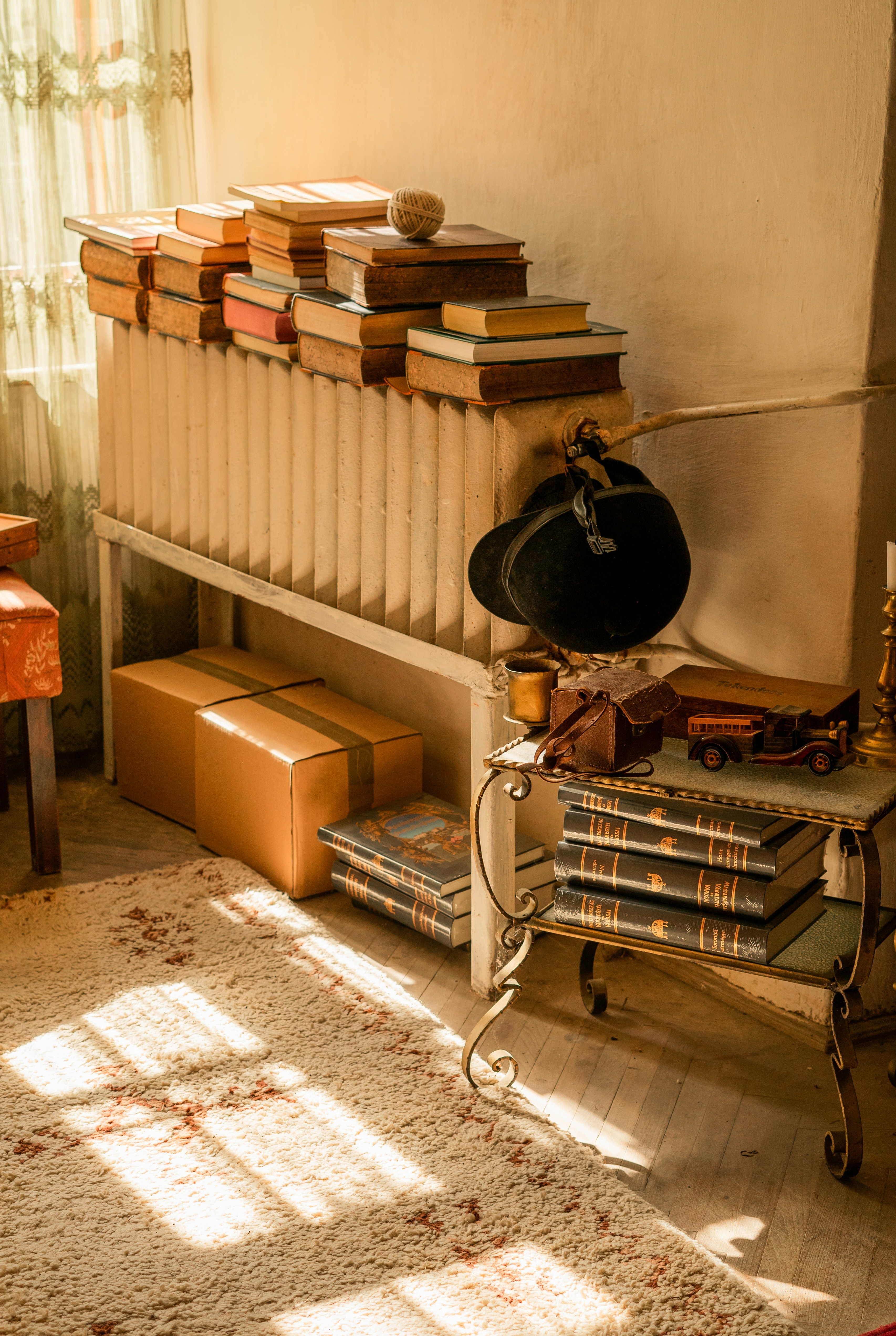 Sunlit interior vignette with a radiator topped by stacked books and a vintage hat. A wheeled table holds more volumes and a toy car, while warm light casts geometric shadows across the rug.
