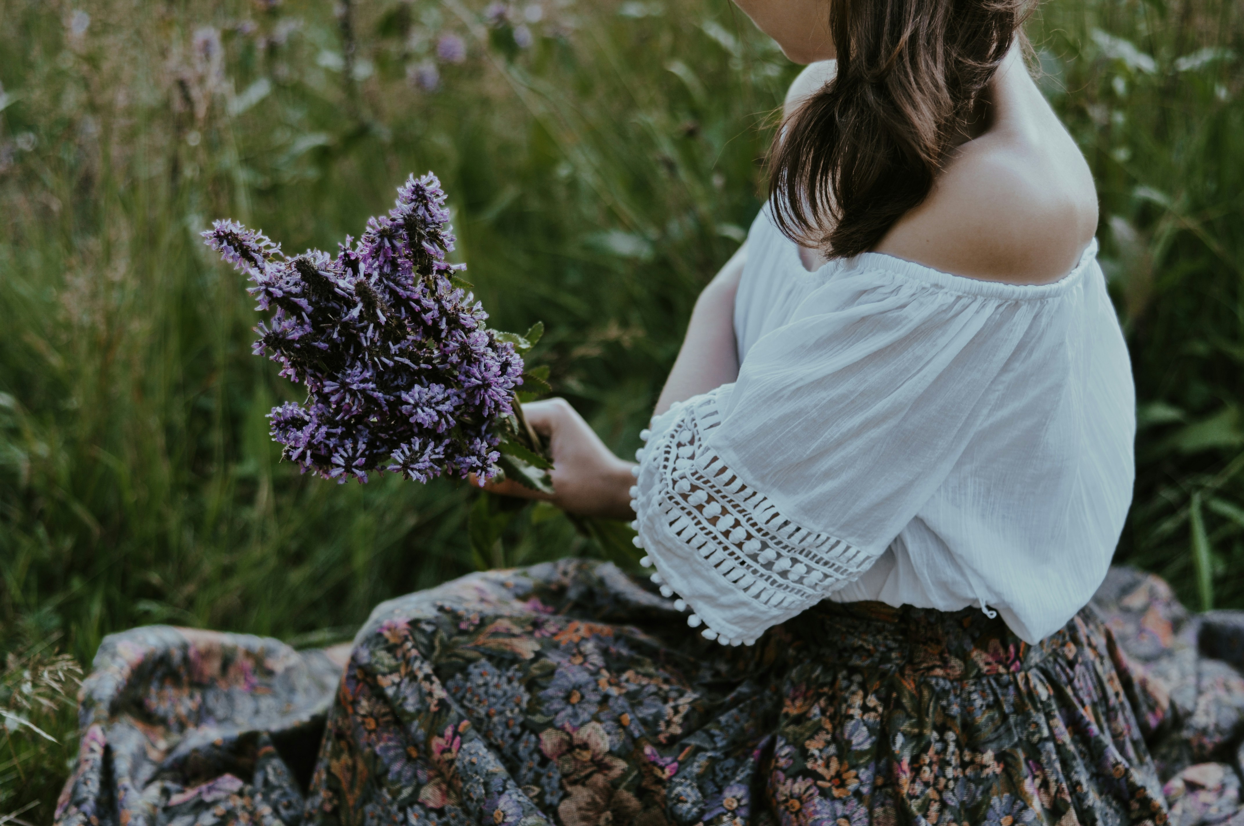 Woman seated in a field, holding a bouquet of purple flowers, dressed in a flowing white top and patterned skirt.