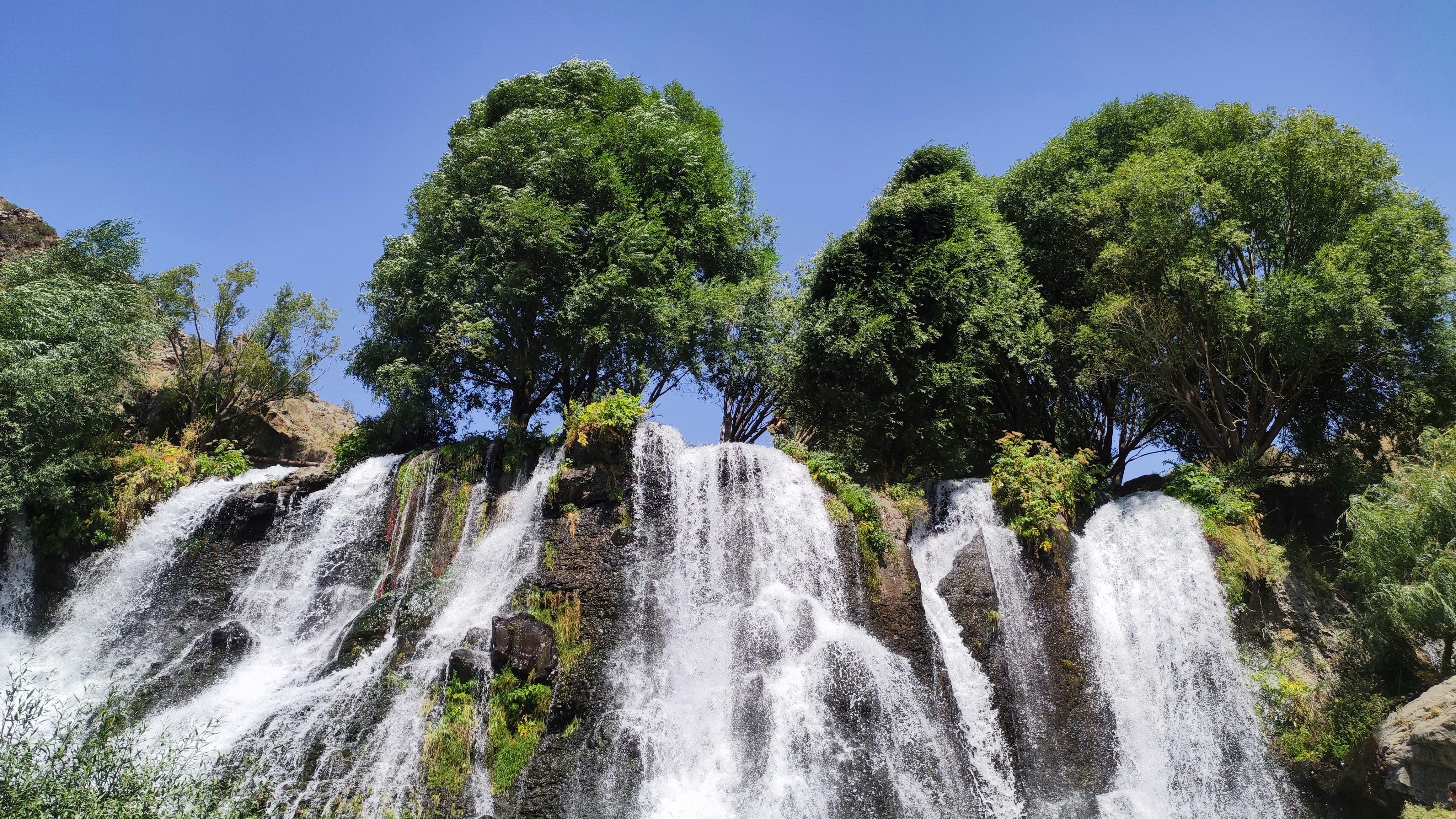 Waterfalls cascading over rocks surrounded by lush green trees under a clear blue sky.