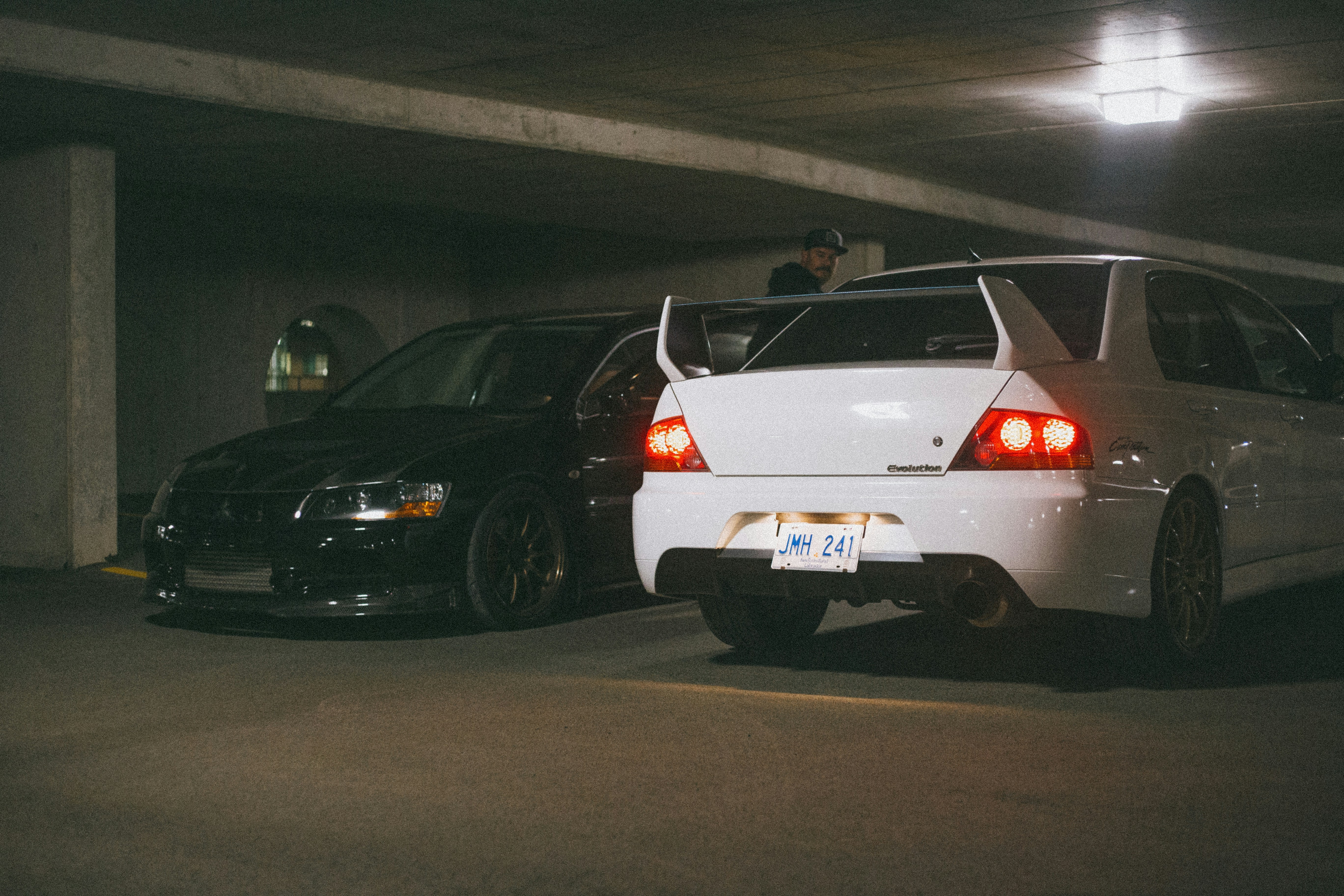 Two modified cars, a black sedan and a white sports car, parked in a dimly lit underground garage, showcasing their sleek designs and custom features.