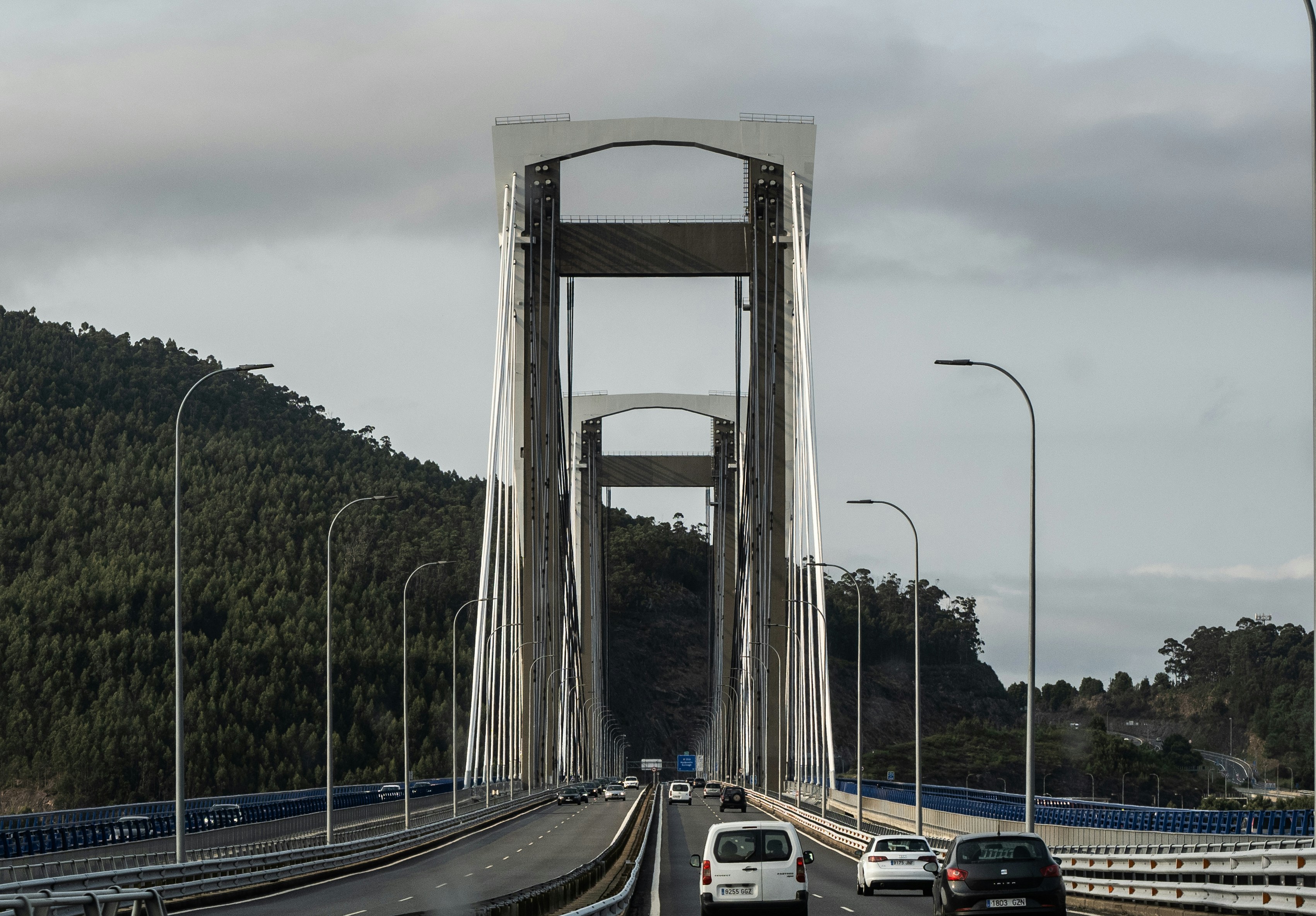 Modern bridge spanning a highway, framed by lush hills under a cloudy sky. Vehicles traverse the structure, highlighting its architectural design.