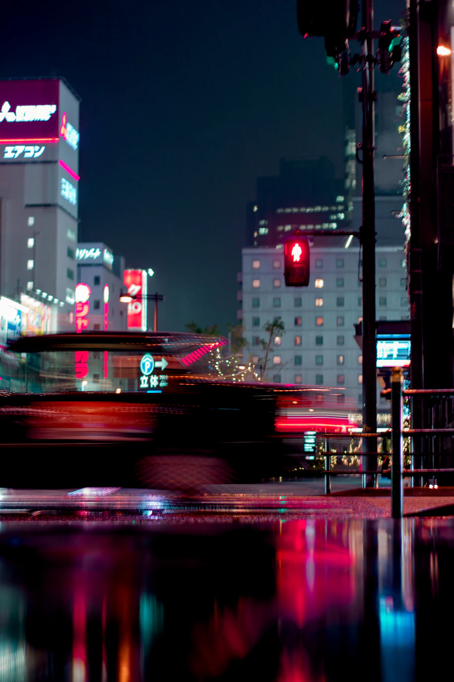 Night city street scene with glowing Japanese signs, a red pedestrian signal, and a fast-moving vehicle streaking across the frame. Reflections of neon lights shimmer on the wet pavement.