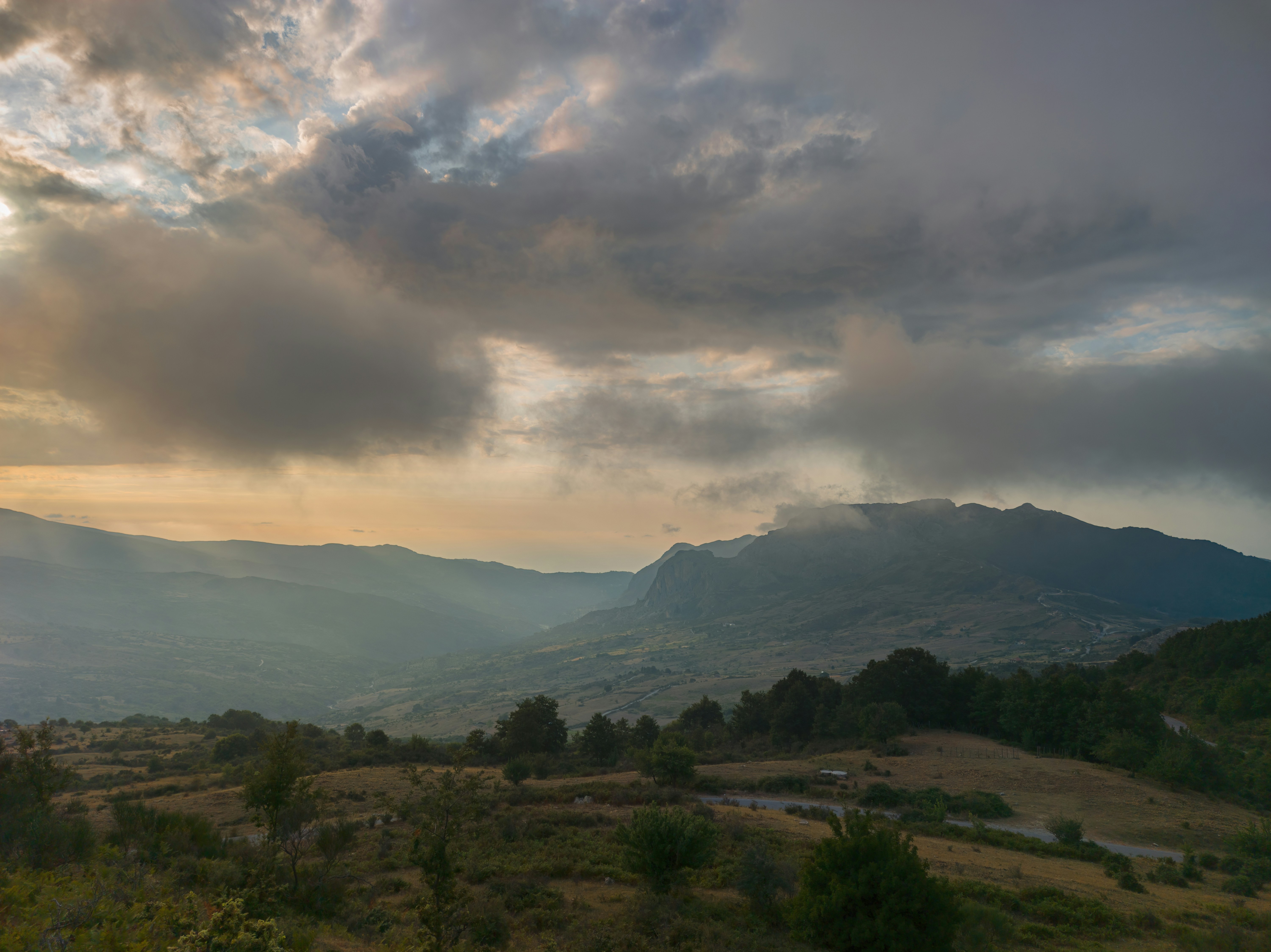 Evening light washes a rolling valley with distant mountains and dramatic clouds, a quiet landscape photograph.