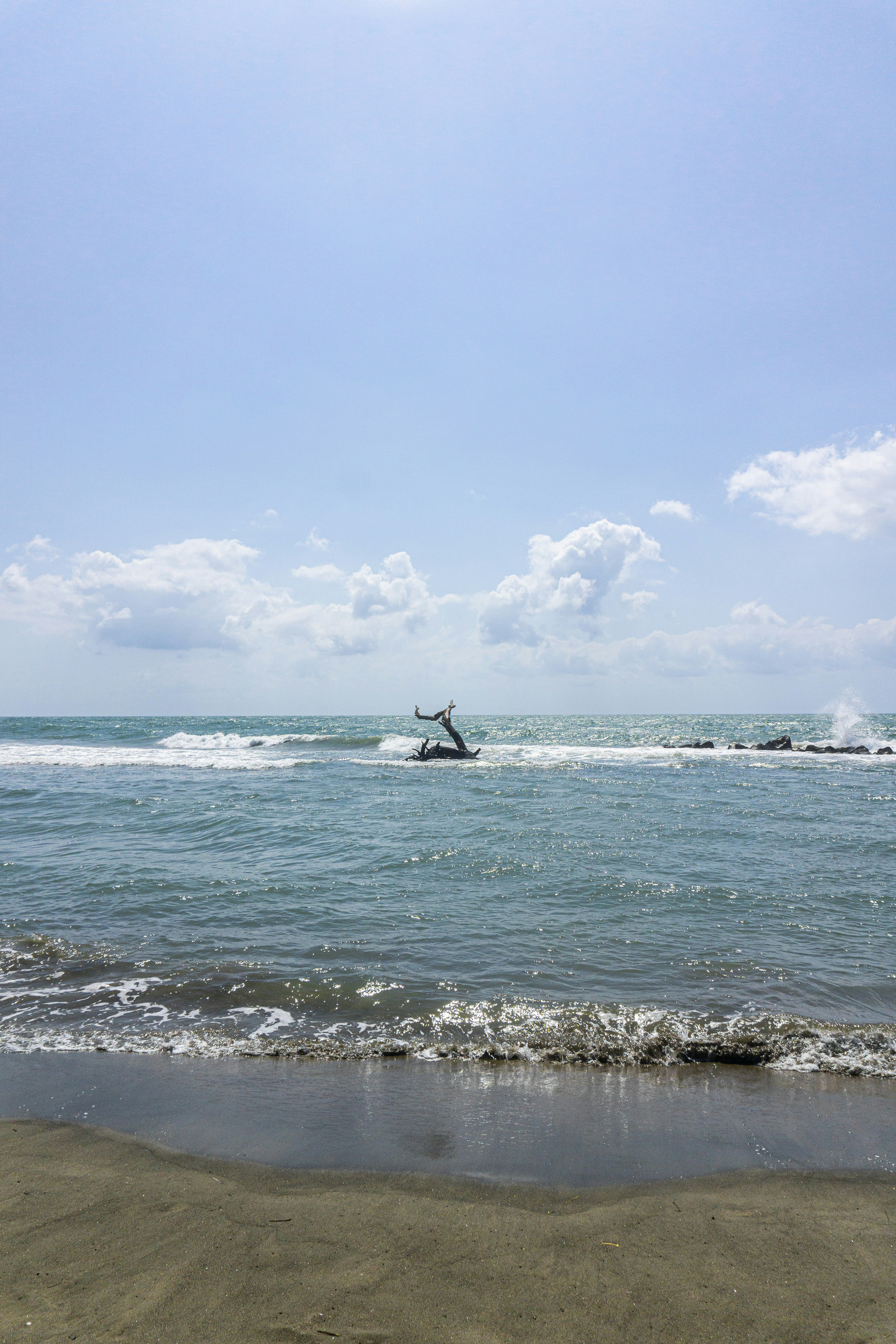 A person balances on a rock amidst gentle ocean waves under a bright sky dotted with clouds.