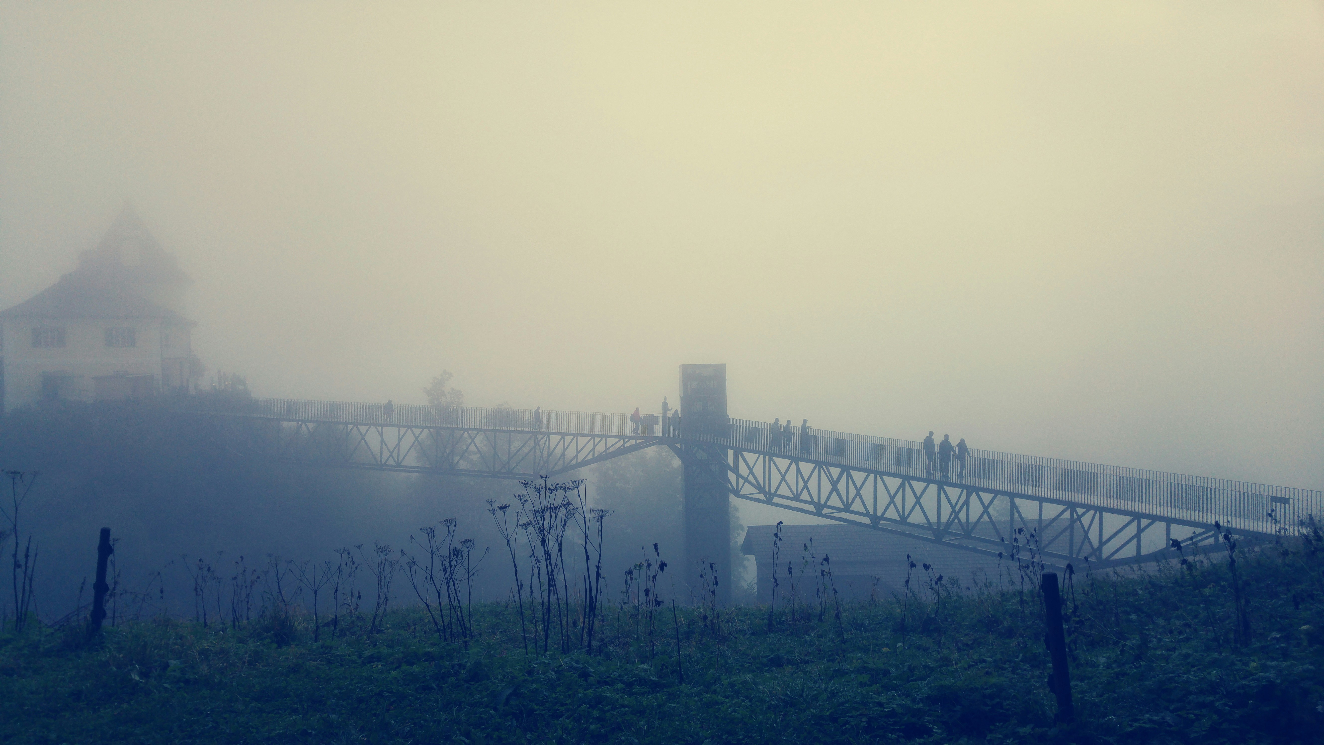 A foggy scene featuring a bridge with silhouettes of people crossing, leading to a distant structure shrouded in mist.