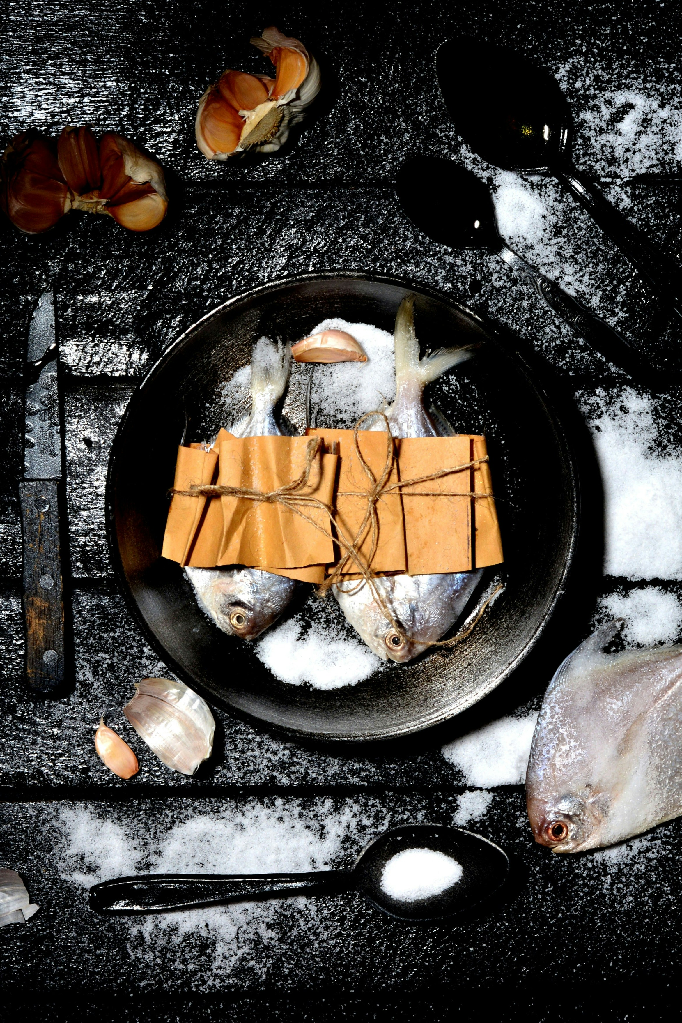 Two small fish bound under parchment in a cast-iron skillet, surrounded by salt and kitchen tools on a dark, textured surface.