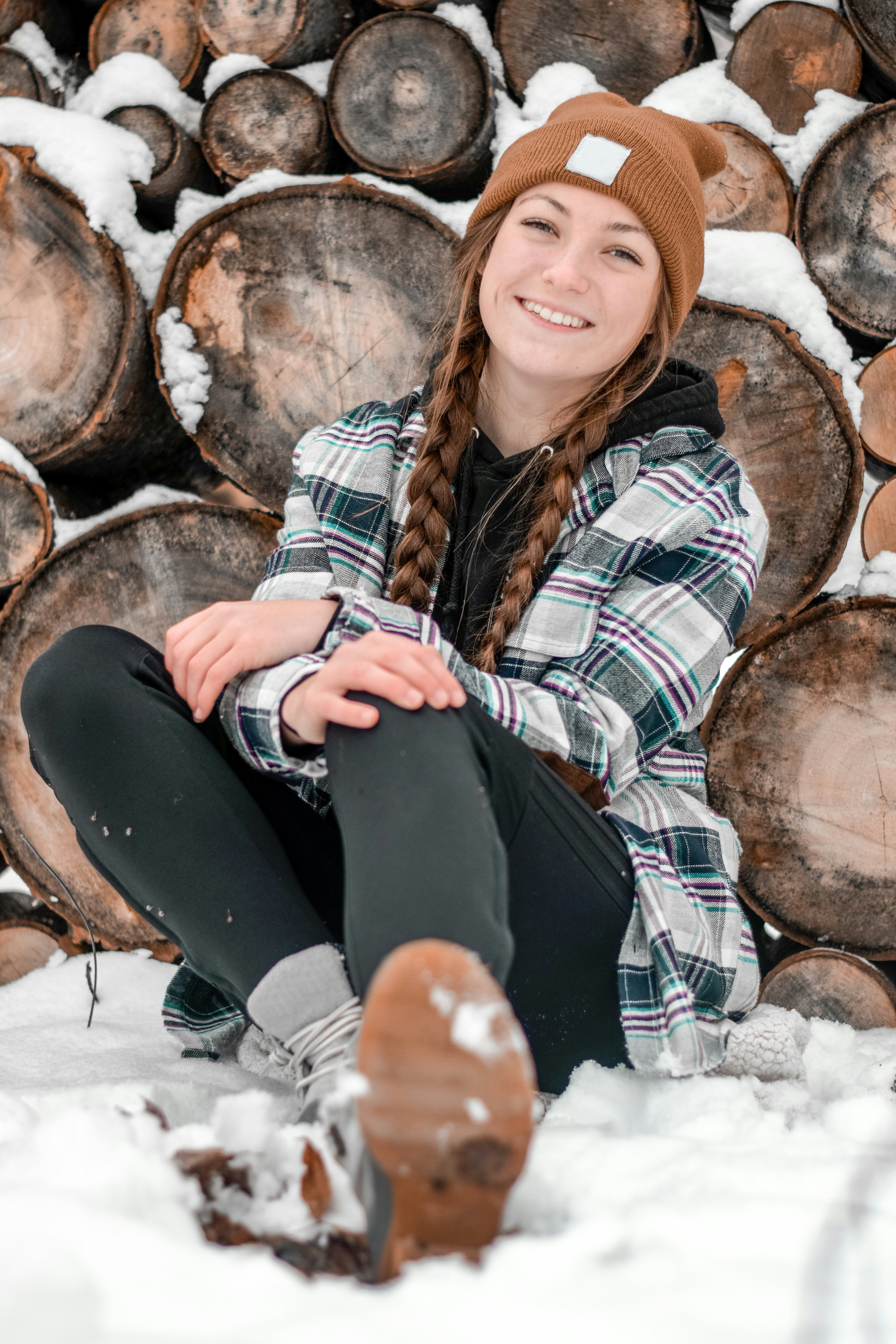 Woman in a plaid jacket and beanie sitting on snow-covered logs, smiling warmly.