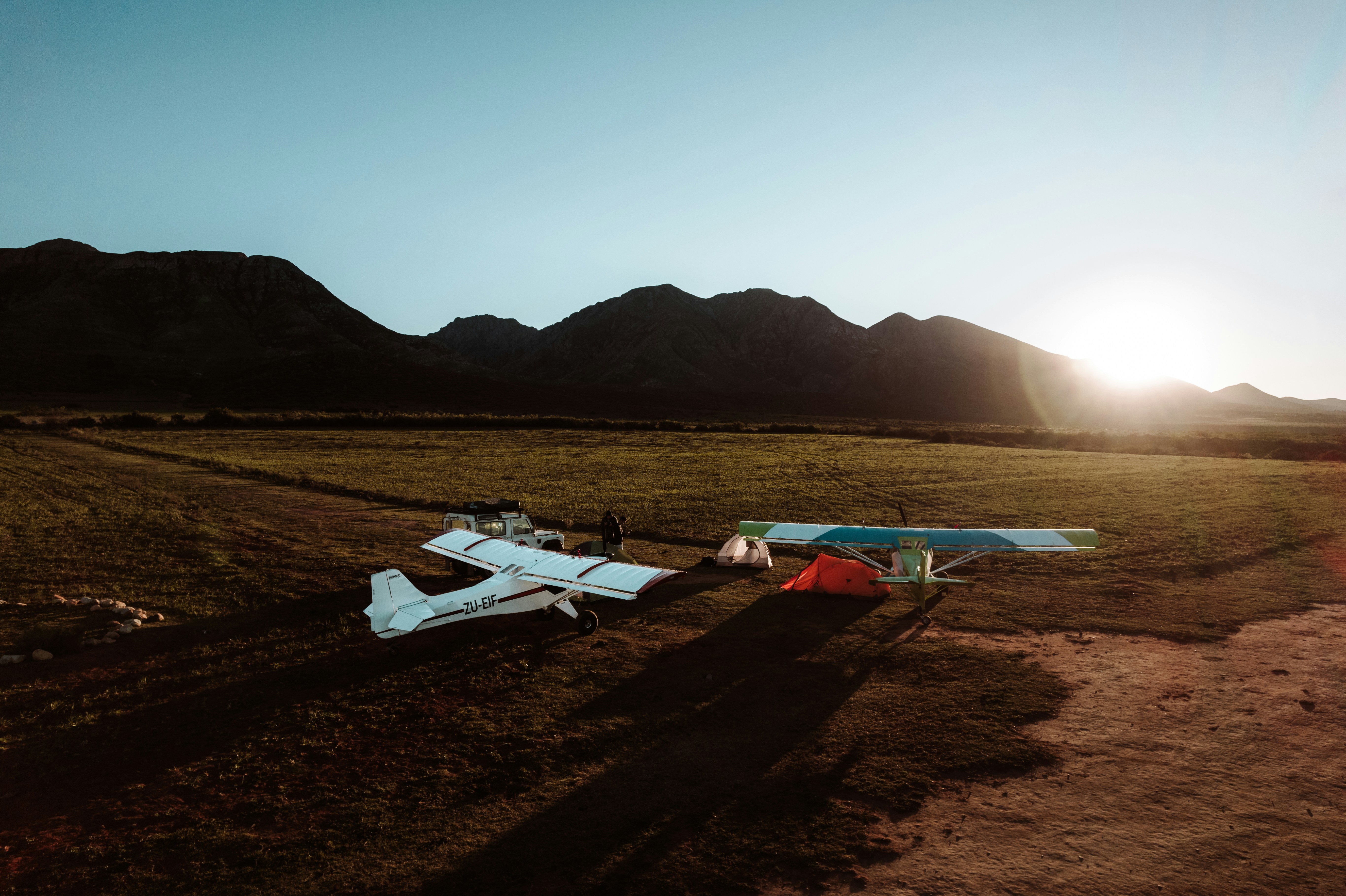 Two small aircraft parked on a grass airstrip at sunset, with mountains silhouetted in the background. A tent is set up nearby, suggesting a spirit of exploration.