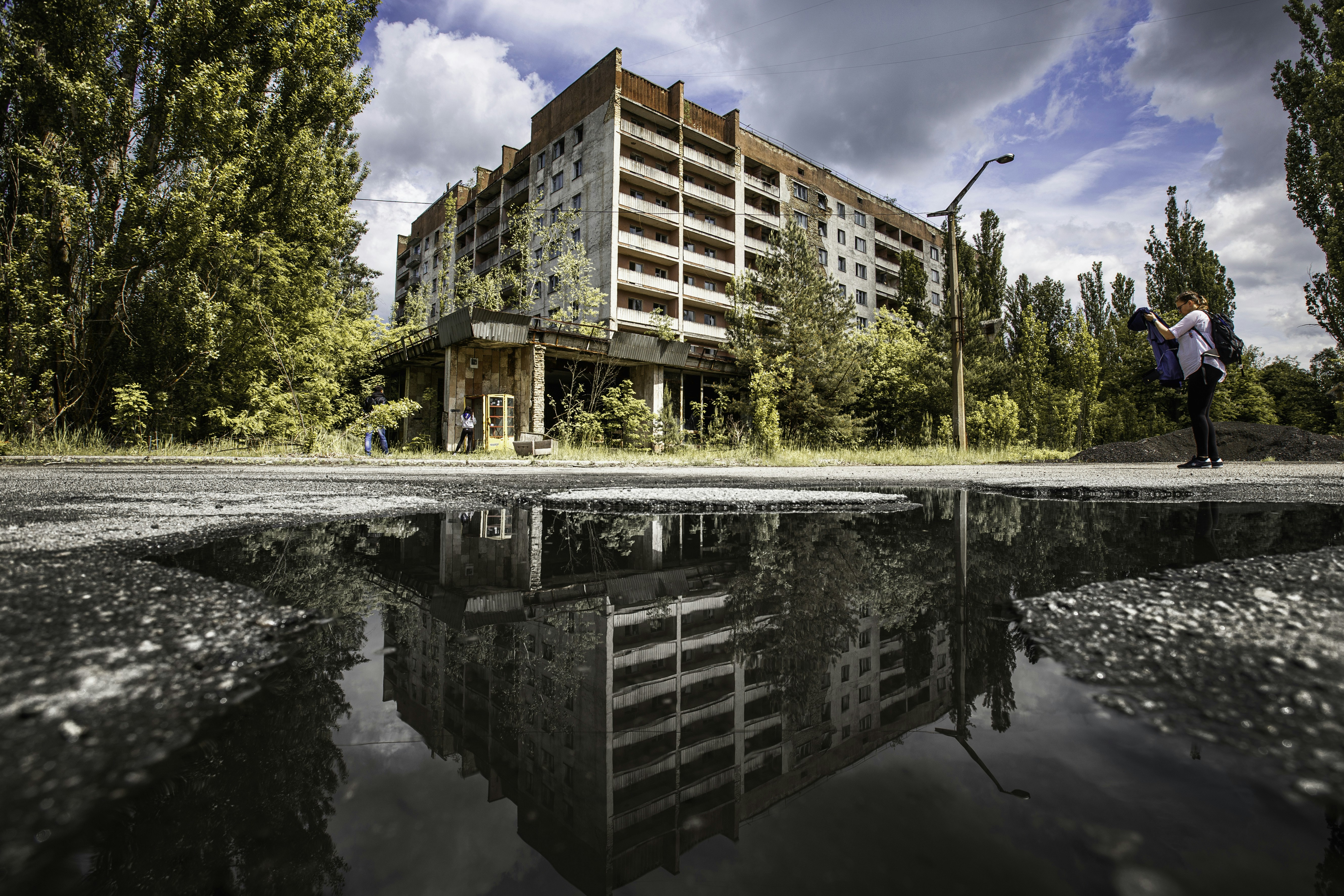 An abandoned building looms over a puddle, reflecting its decaying structure amidst overgrown greenery. Two figures stand nearby, adding a sense of scale to the haunting scene.