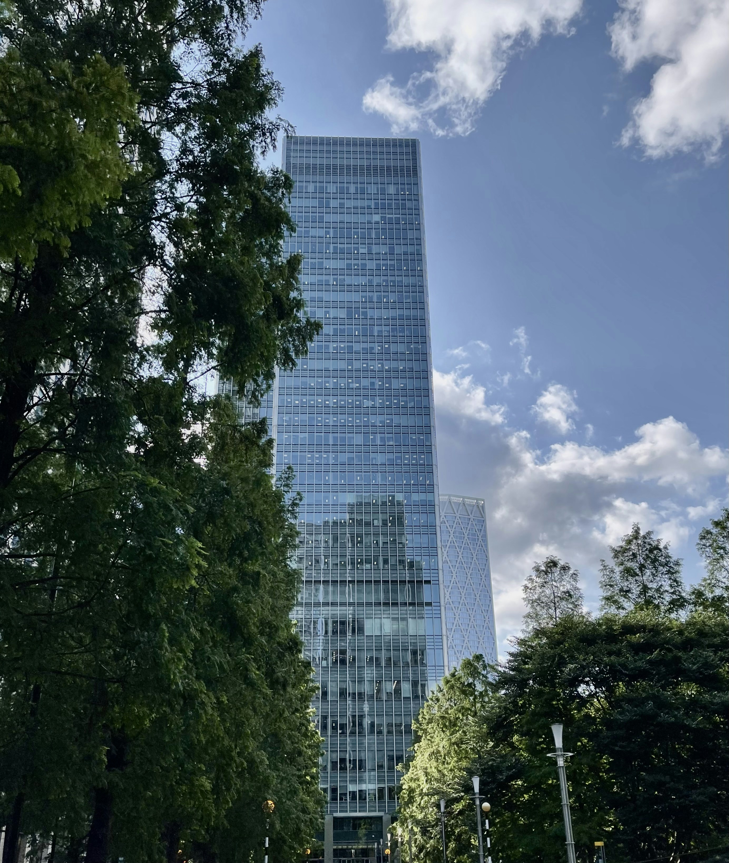 A sleek glass skyscraper rises amidst lush greenery, showcasing the harmony between urban architecture and nature. Fluffy clouds drift in a bright blue sky.