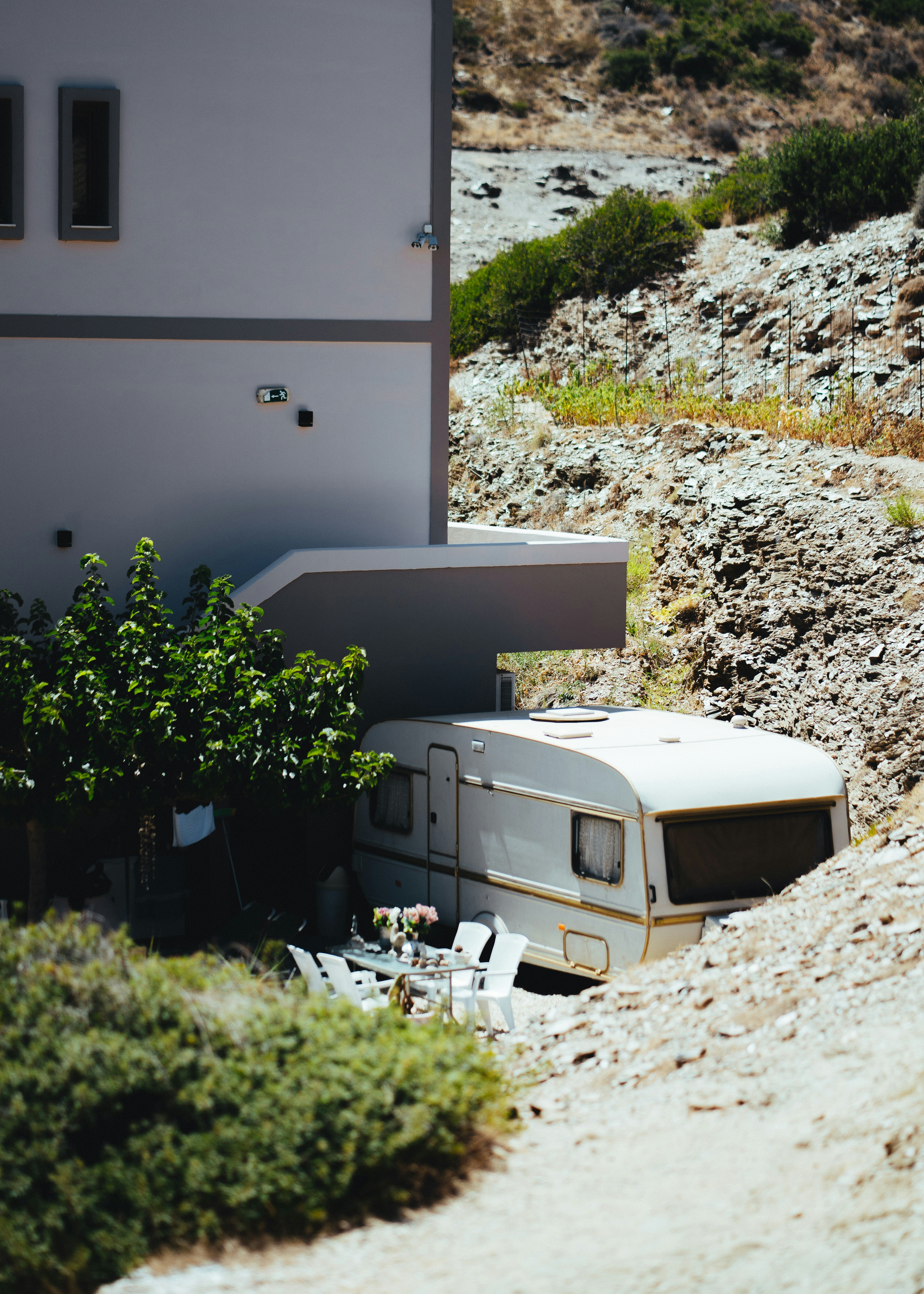 A cozy caravan nestled beside a modern building, surrounded by lush greenery and a tranquil outdoor seating area. The scene captures a harmonious blend of nature and architecture.