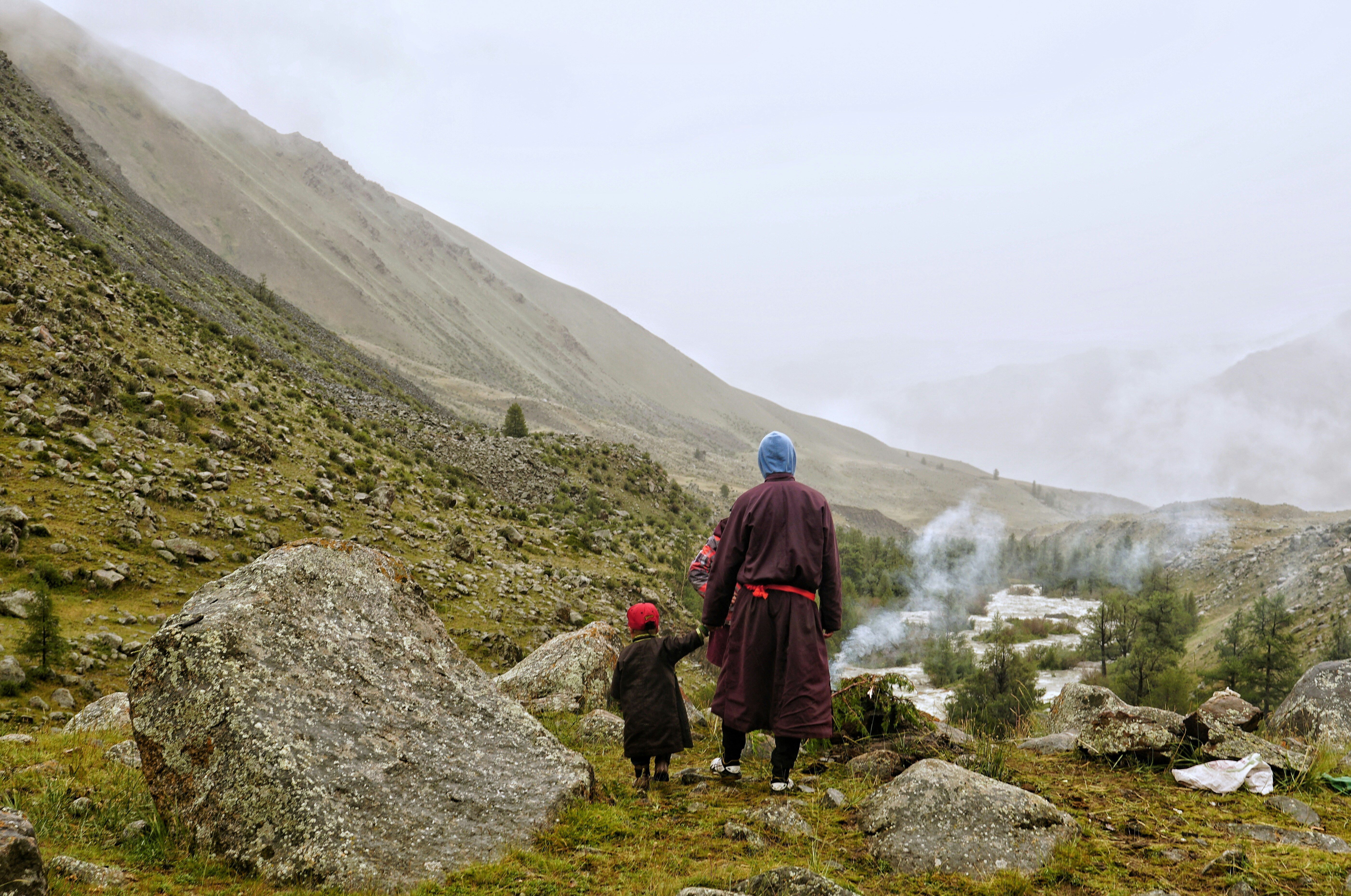Two figures in a misty highland pass lead the way among boulders and scrub