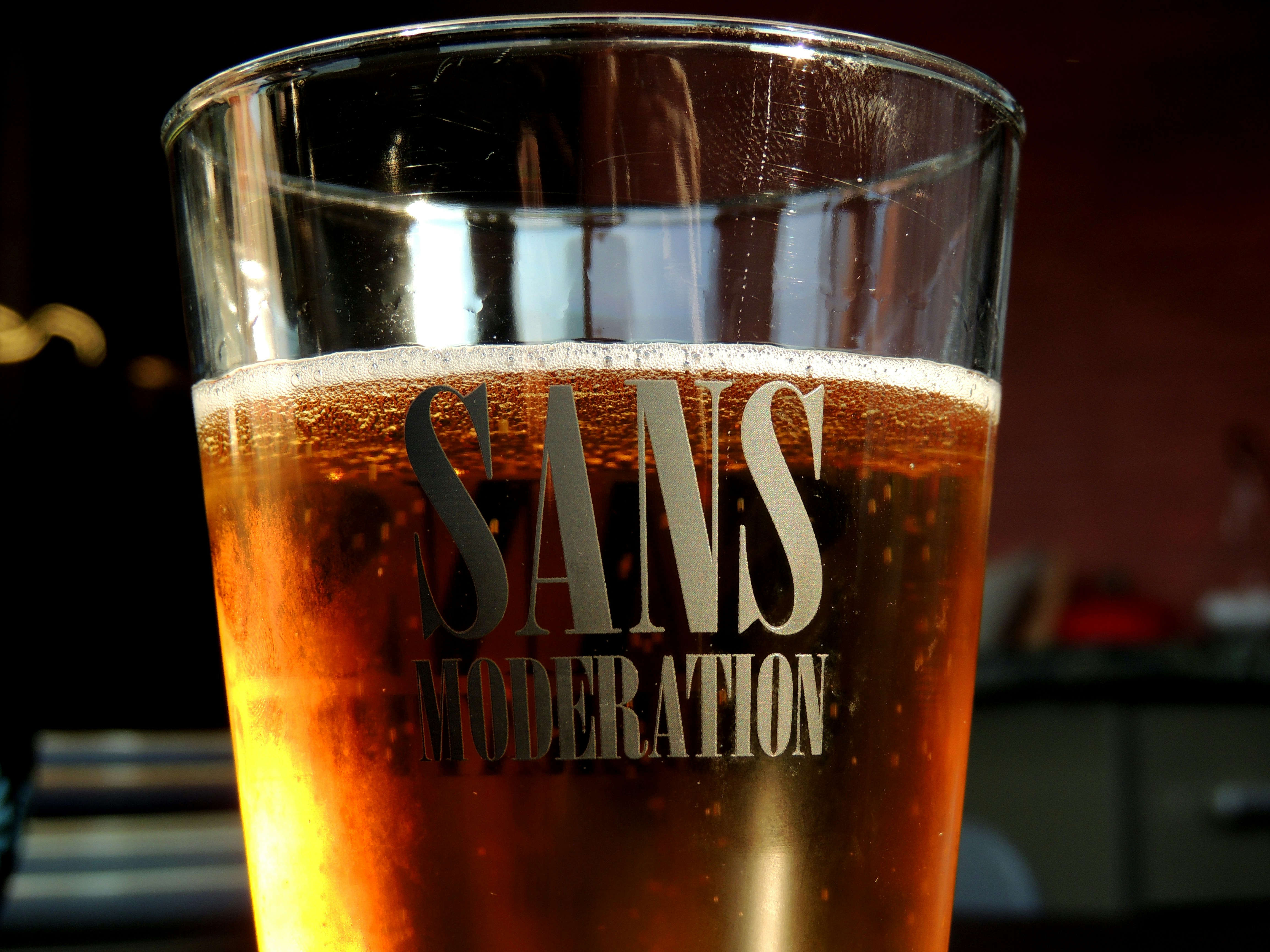 Close-up of a beer glass with 'SANS MODERATION' text, filled with amber liquid reflecting warm light.
