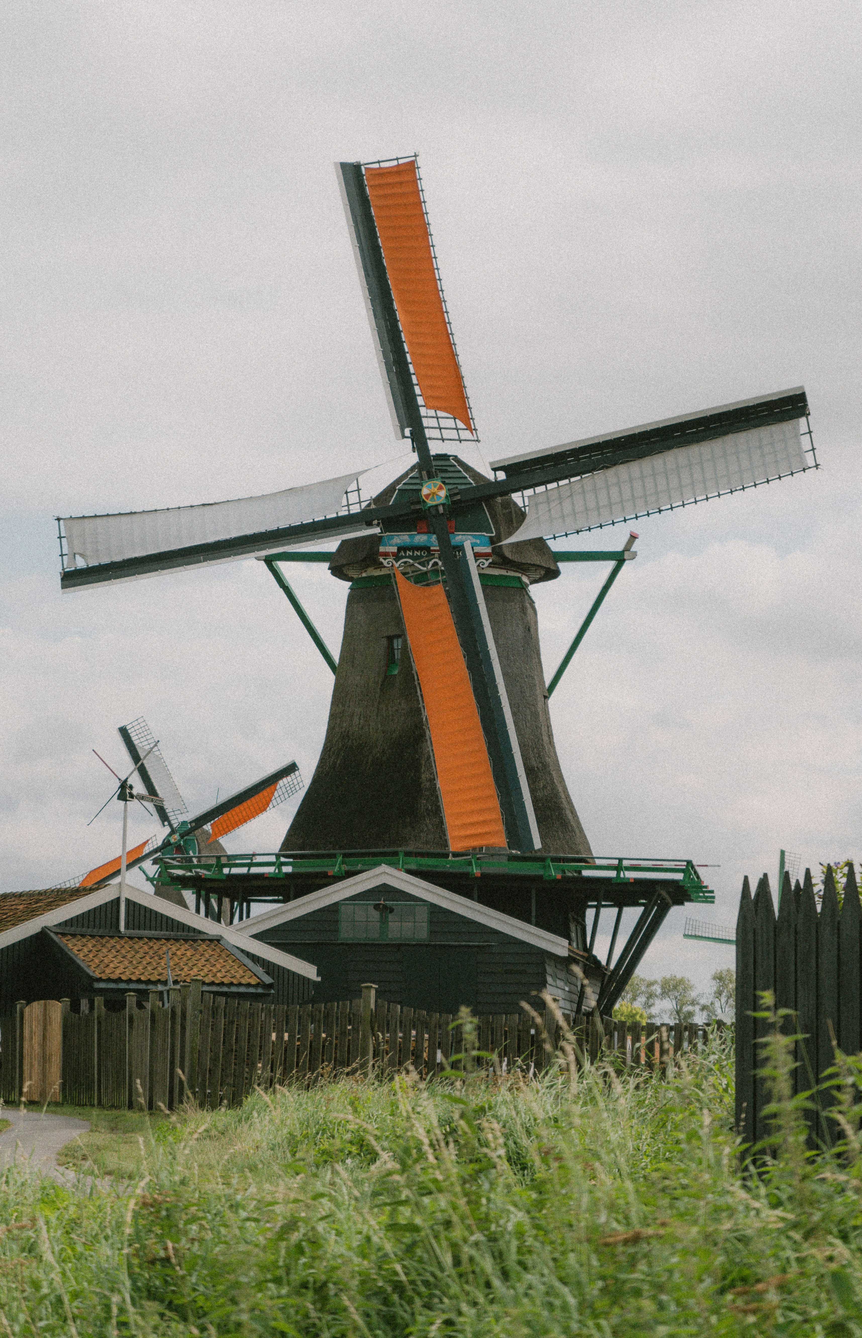 A traditional Dutch windmill stands tall against a cloudy sky, its vibrant orange sails contrasting with the rustic surroundings. The scene captures the essence of rural heritage.