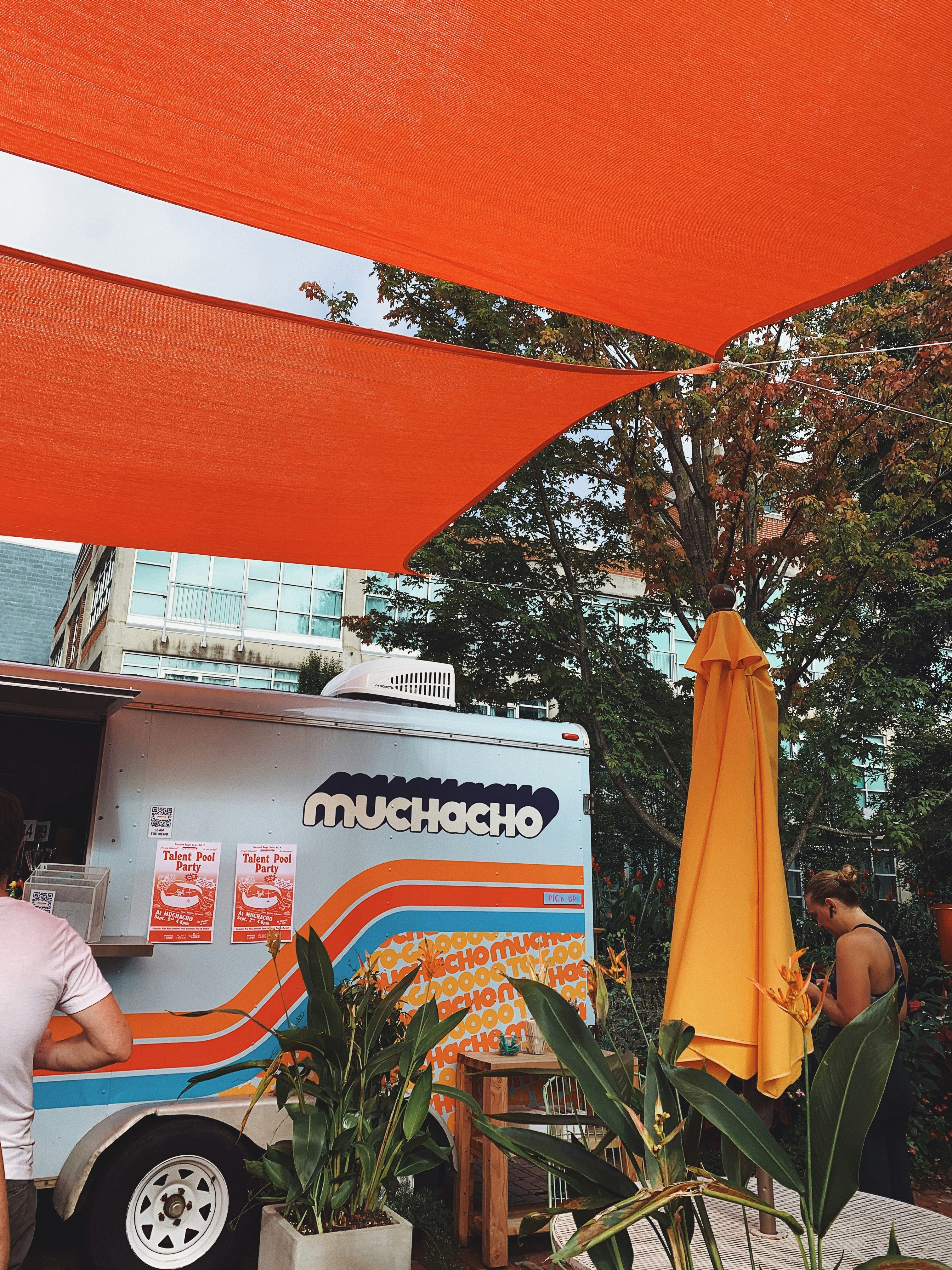 Vibrant food truck named Muchacho nestled among lush greenery and colorful canopies, inviting patrons to enjoy outdoor dining.