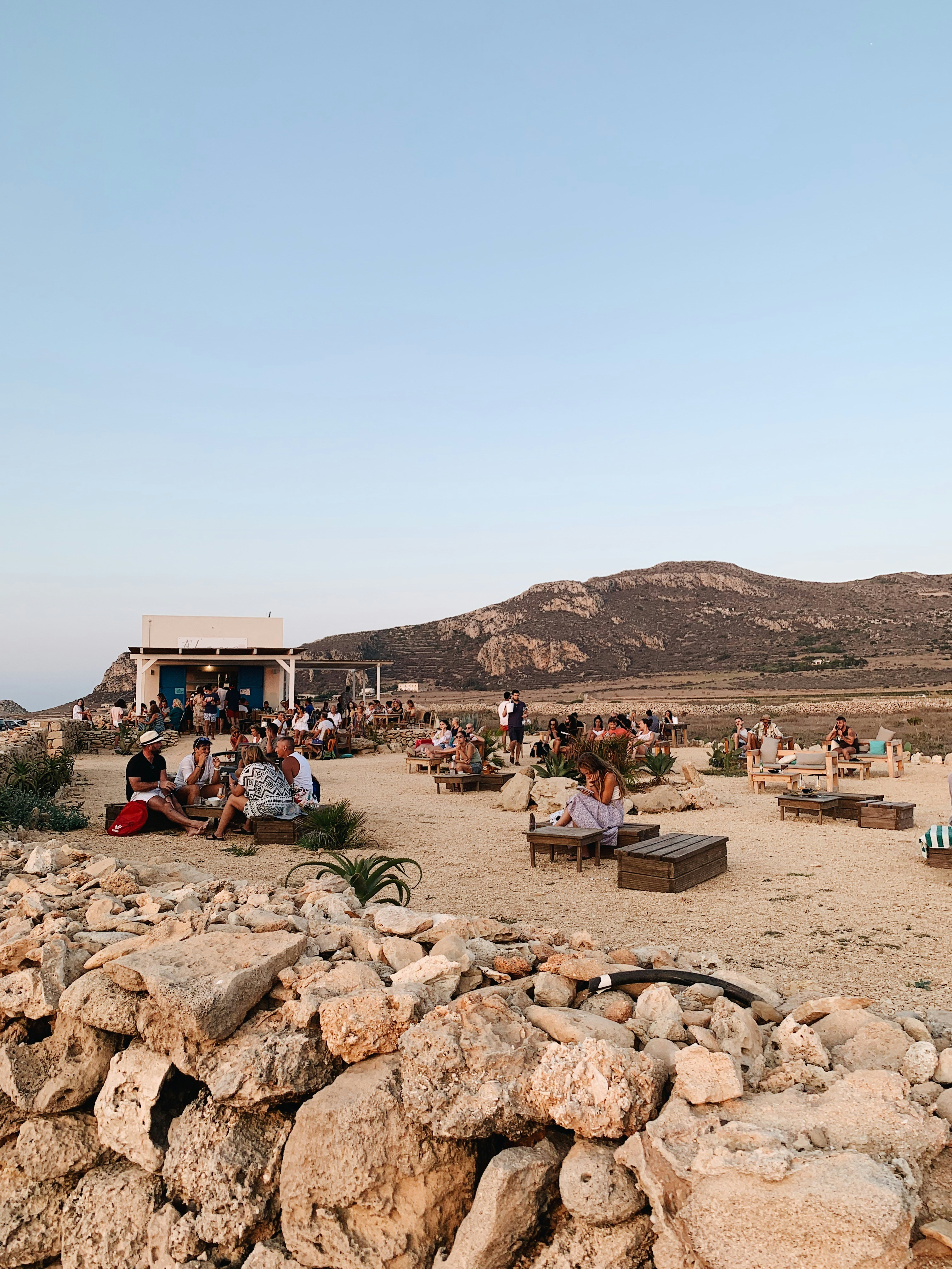 Outdoor gathering space filled with people enjoying a leisurely evening, surrounded by rock formations and a scenic backdrop. The atmosphere is relaxed and inviting.