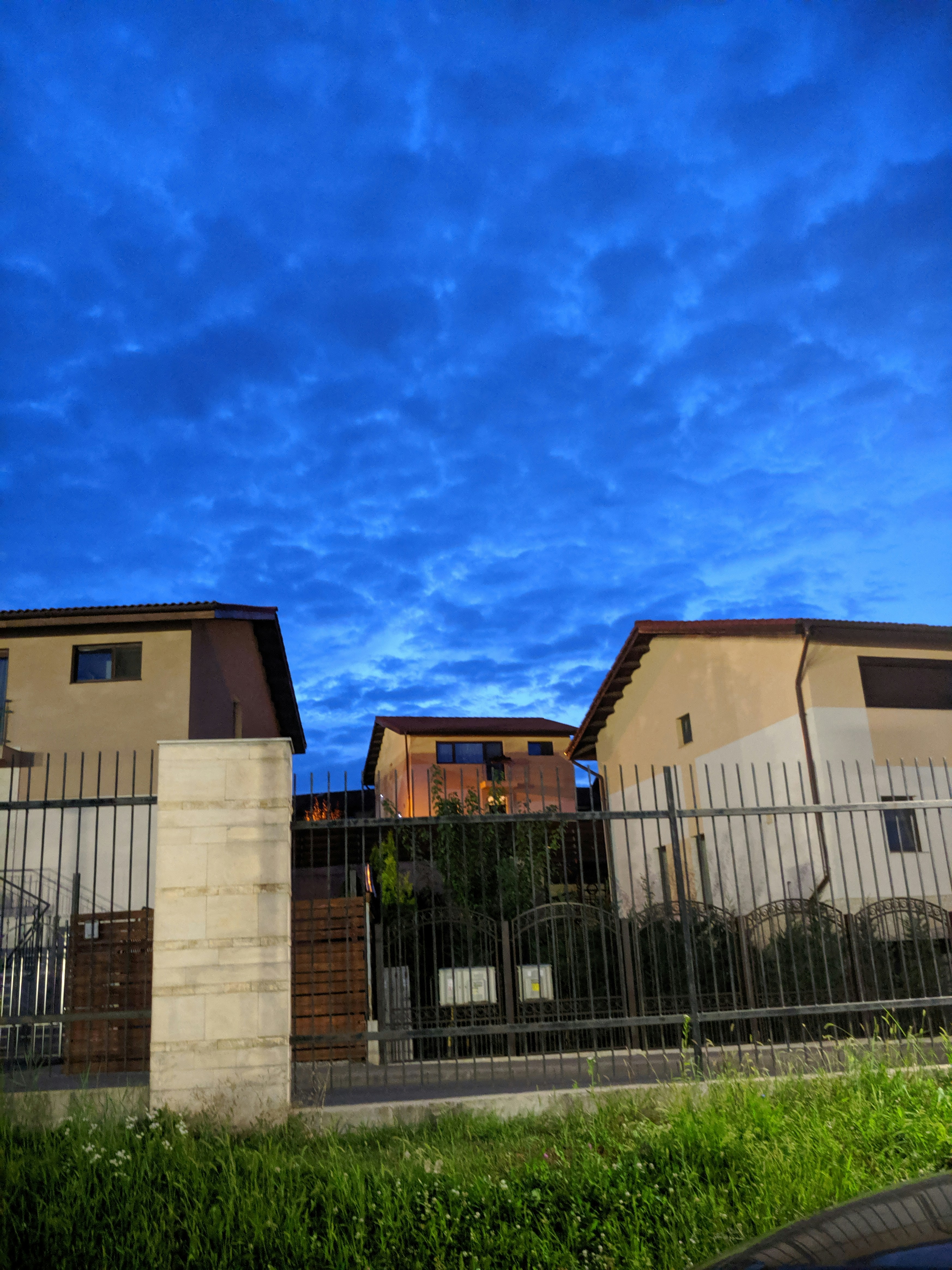 Bright blue sky with wispy clouds above a row of beige houses behind a metal fence. A grassy verge frames the foreground.