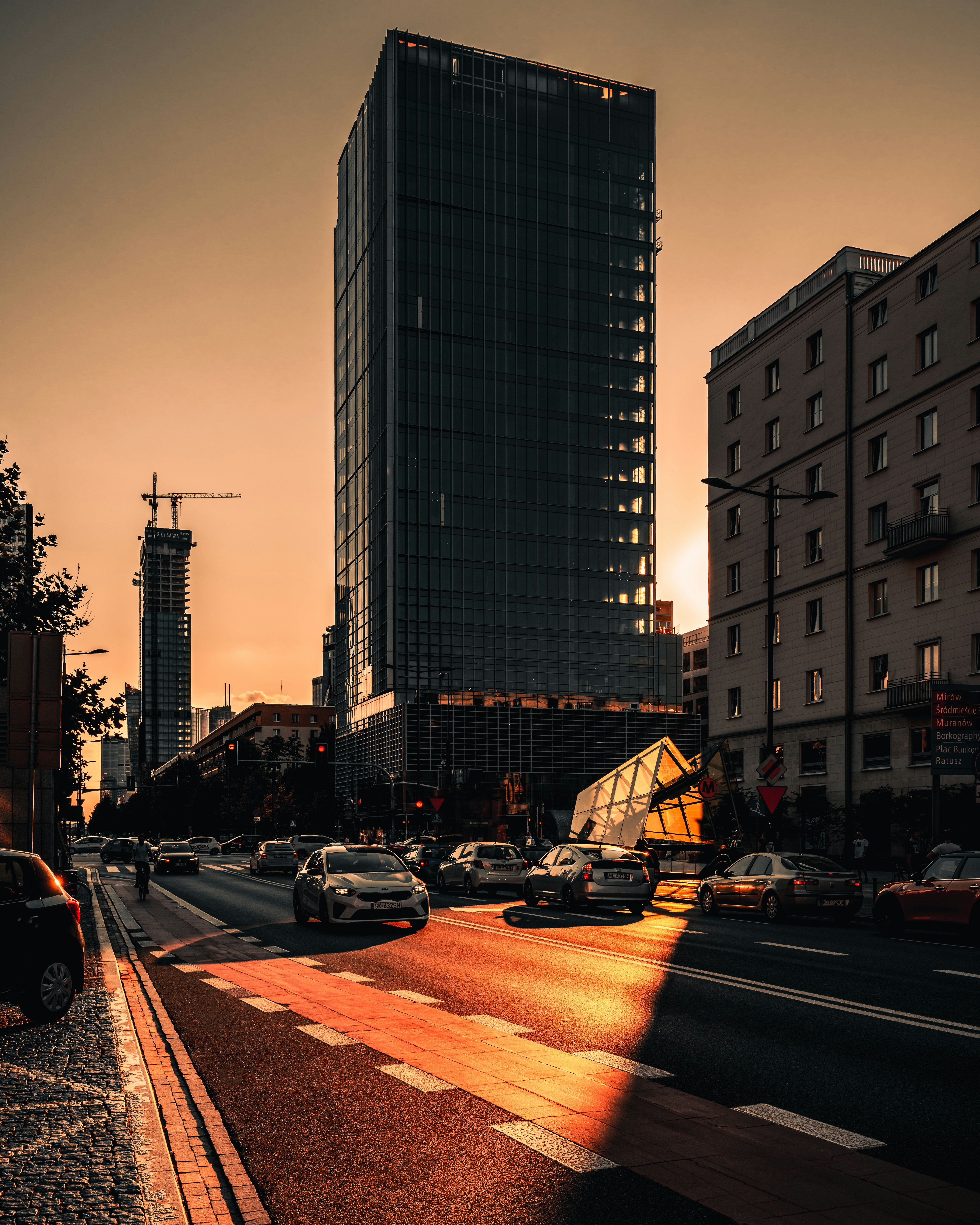 Modern skyscrapers silhouetted against a golden sunset, with bustling traffic and pedestrians below. The scene captures the essence of urban life transitioning into evening.