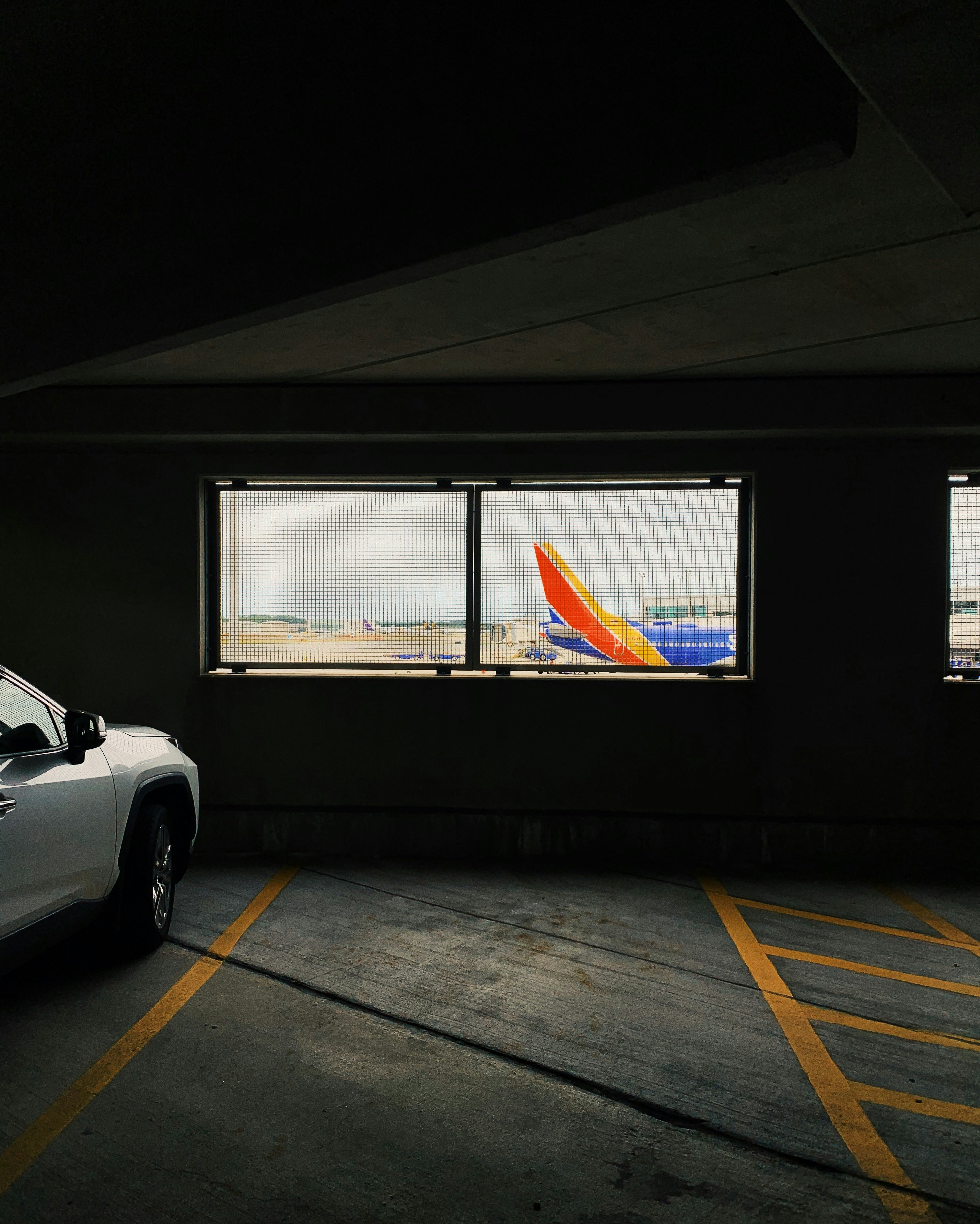White SUV parked in a dimly lit garage, framed by a window revealing colorful airline tails in the background. 
