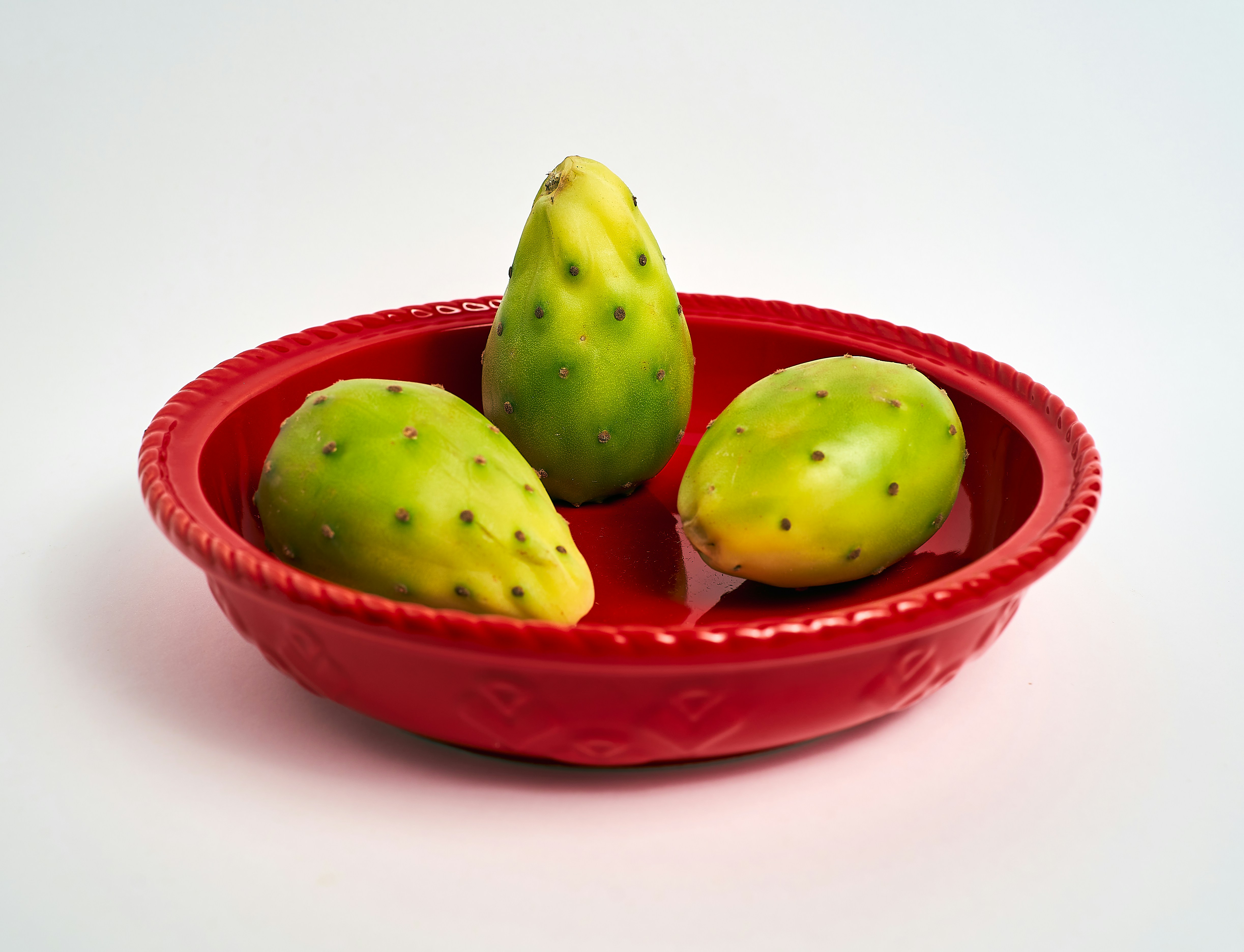 Three prickly pears in a red ceramic bowl on a clean white surface