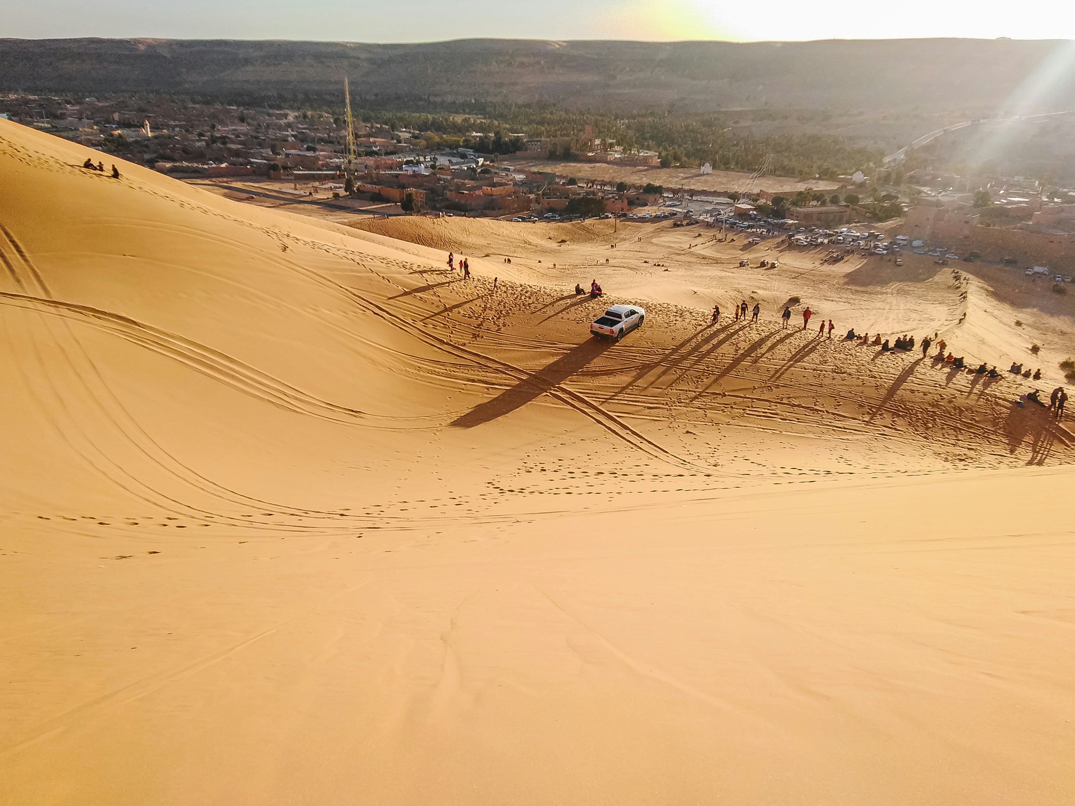 A white vehicle navigates the golden sands of a vast desert, with people enjoying the landscape in the background. The scene captures the blend of adventure and natural beauty.