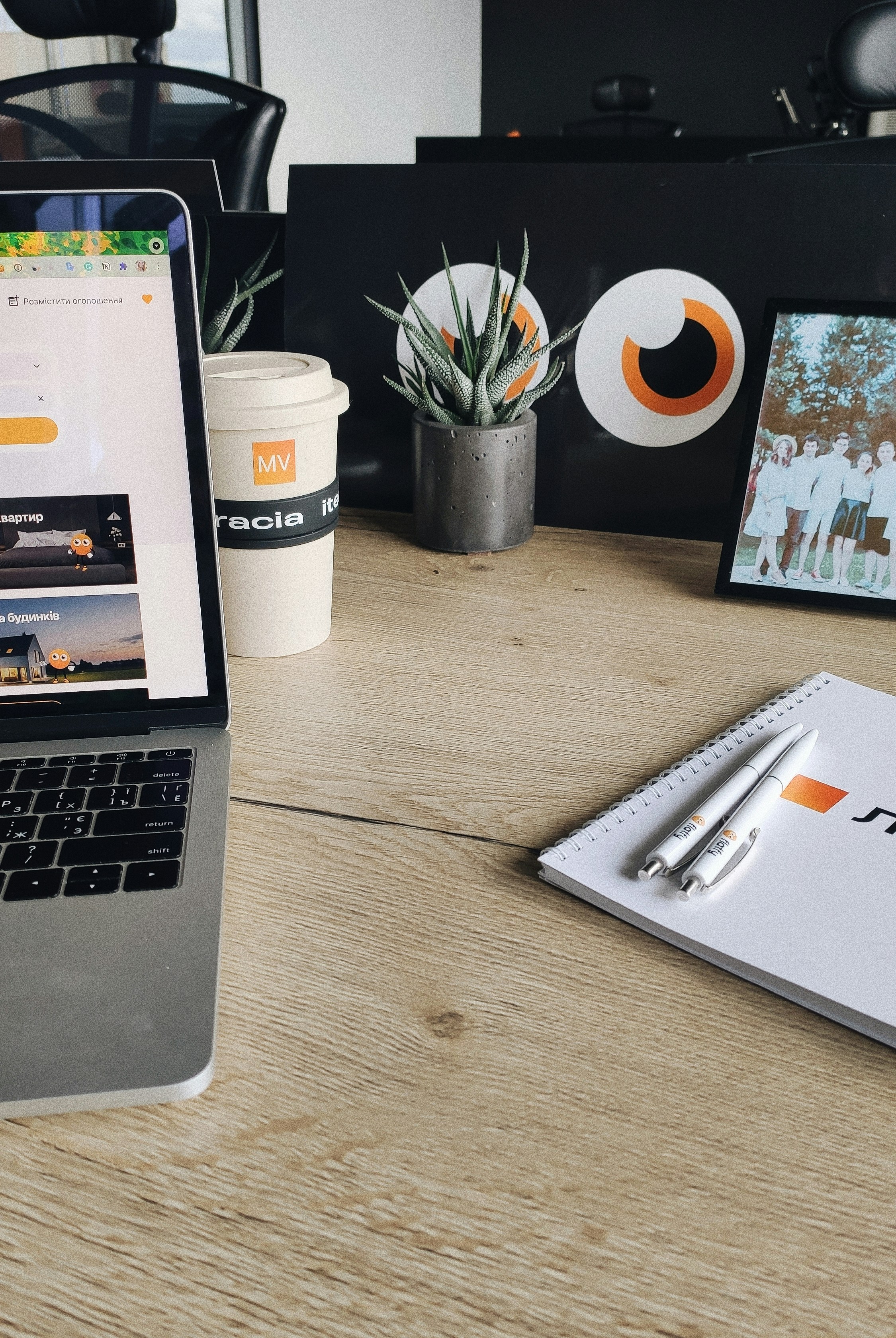 A tidy desk scene with a laptop on the left, a white takeaway cup, a small succulent in a gray pot, framed photos, and a notebook with pens on a wooden surface.