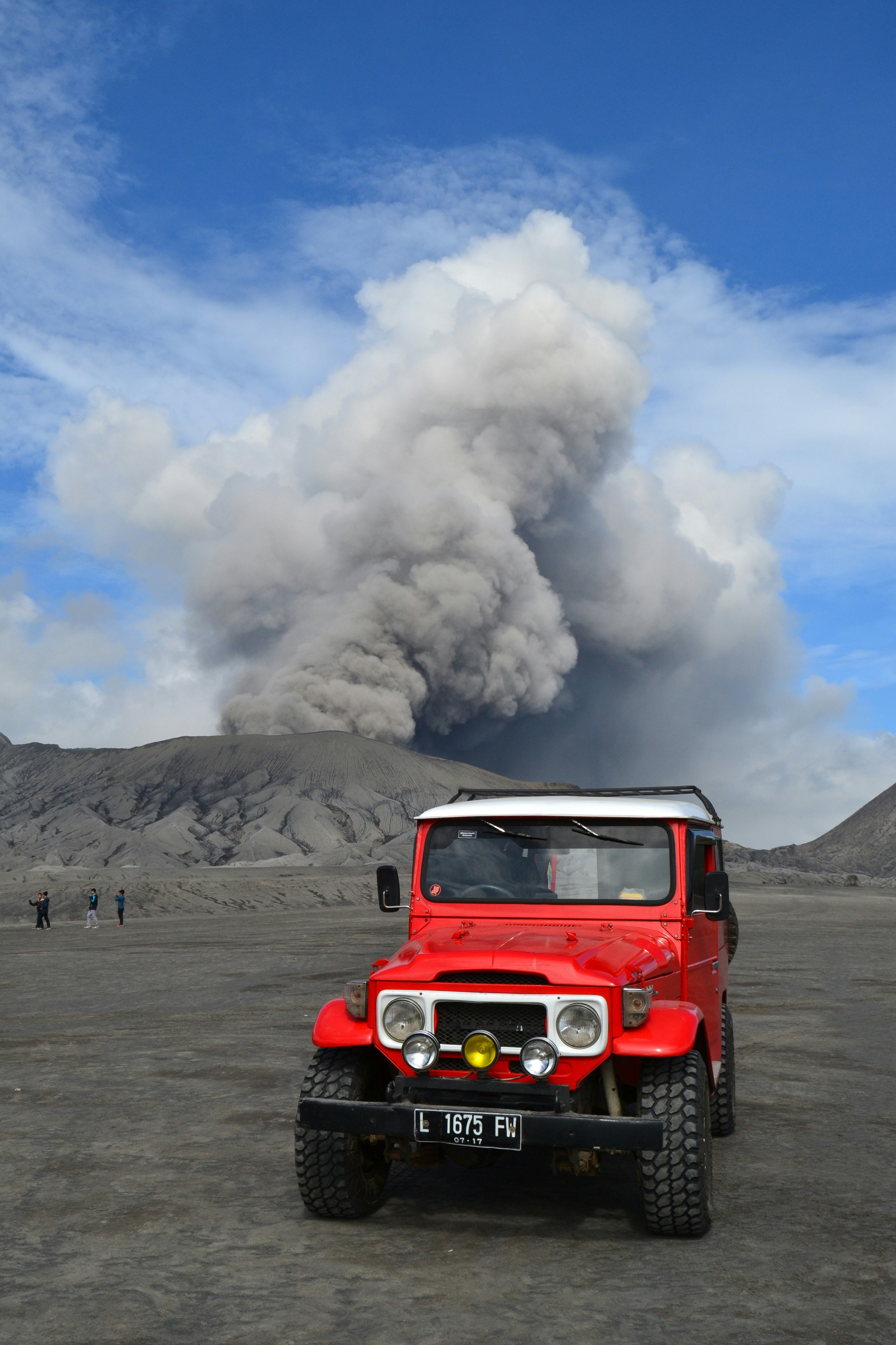 Bright red off-road vehicle parked on volcanic terrain with an erupting volcano in the background, showcasing the contrast between man-made and natural power.