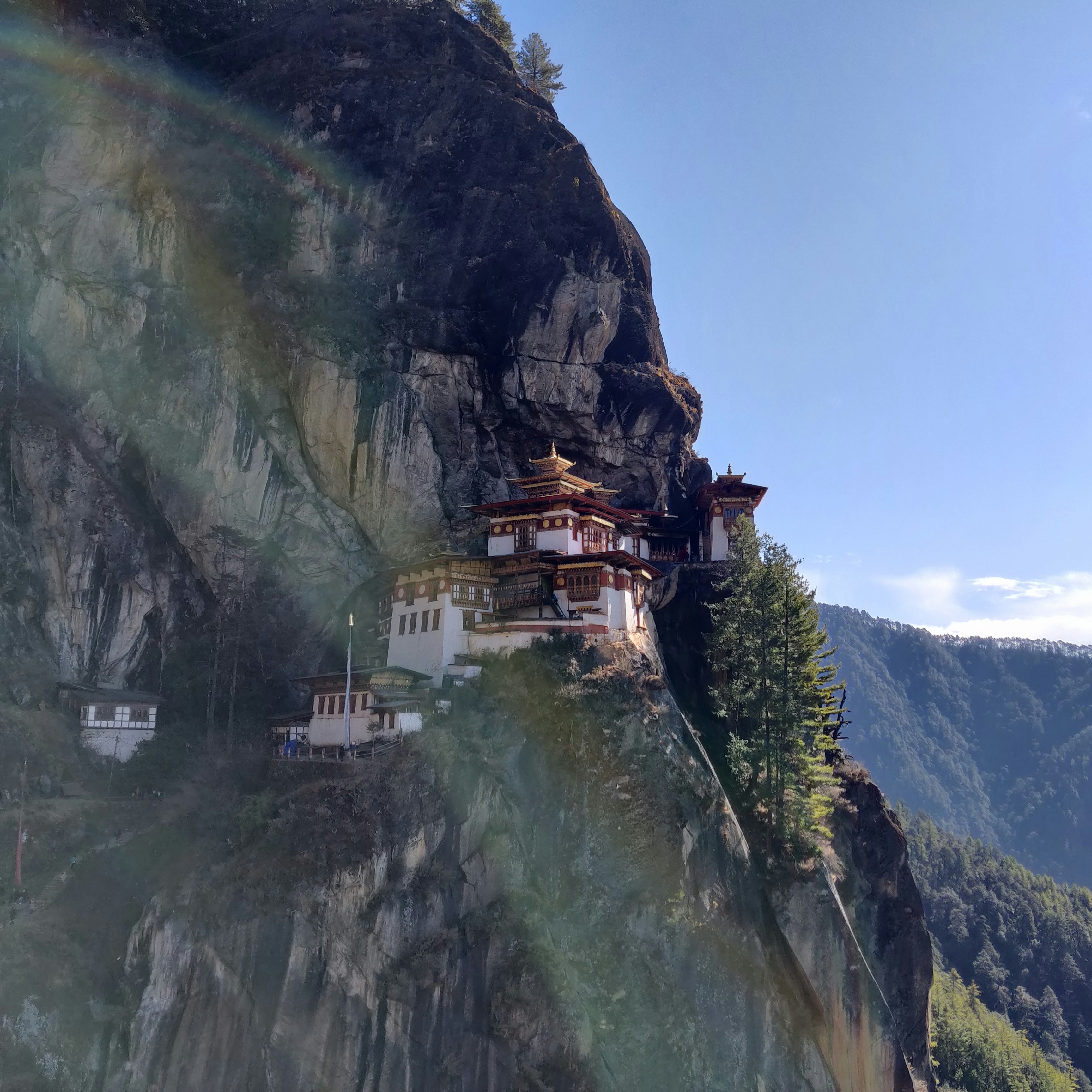 A majestic monastery nestled on a cliffside, surrounded by lush greenery and towering mountains under a clear blue sky.