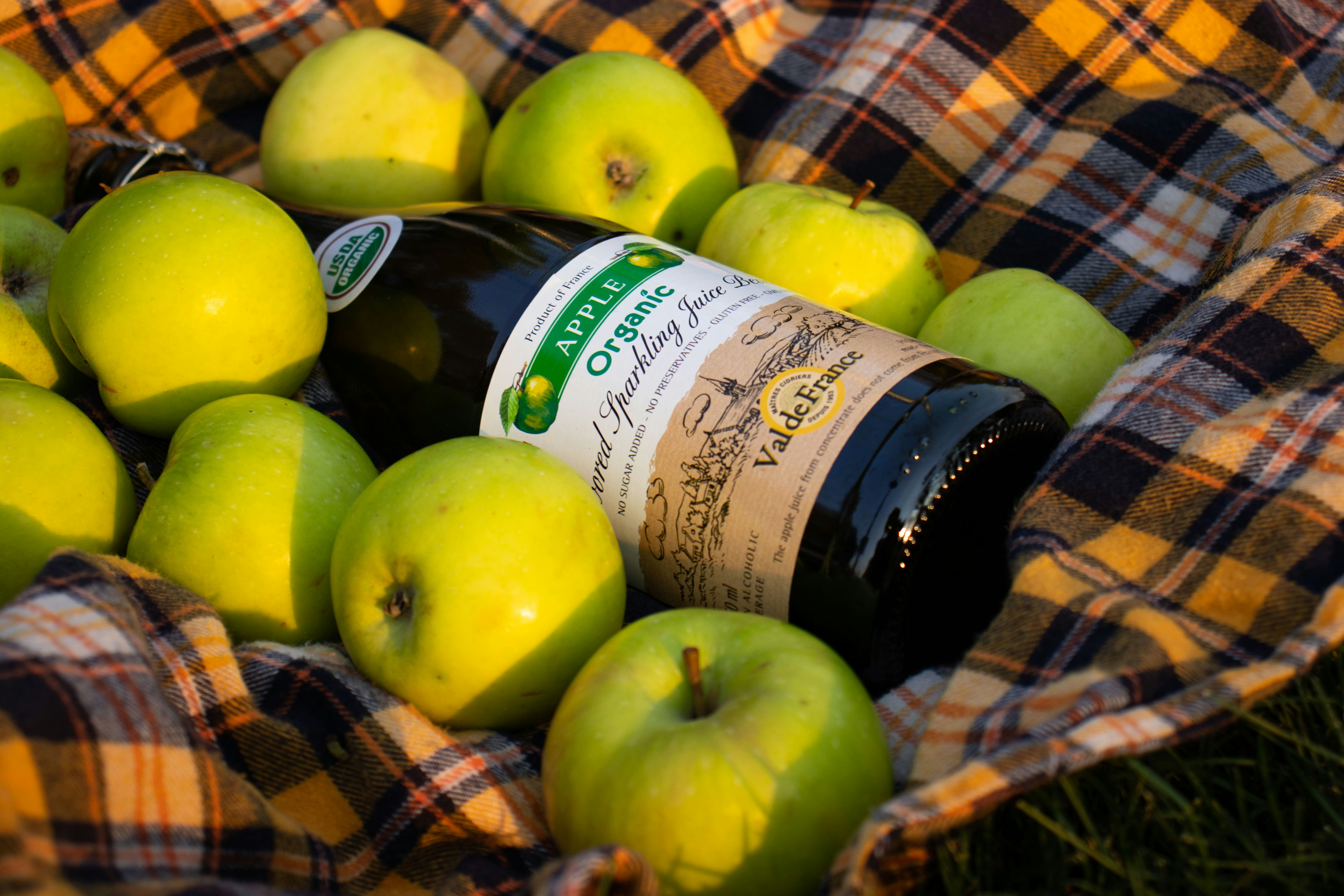 A bottle of organic apple sparkling juice nestled among vibrant green apples on a checkered blanket. The scene evokes a sense of autumnal abundance.