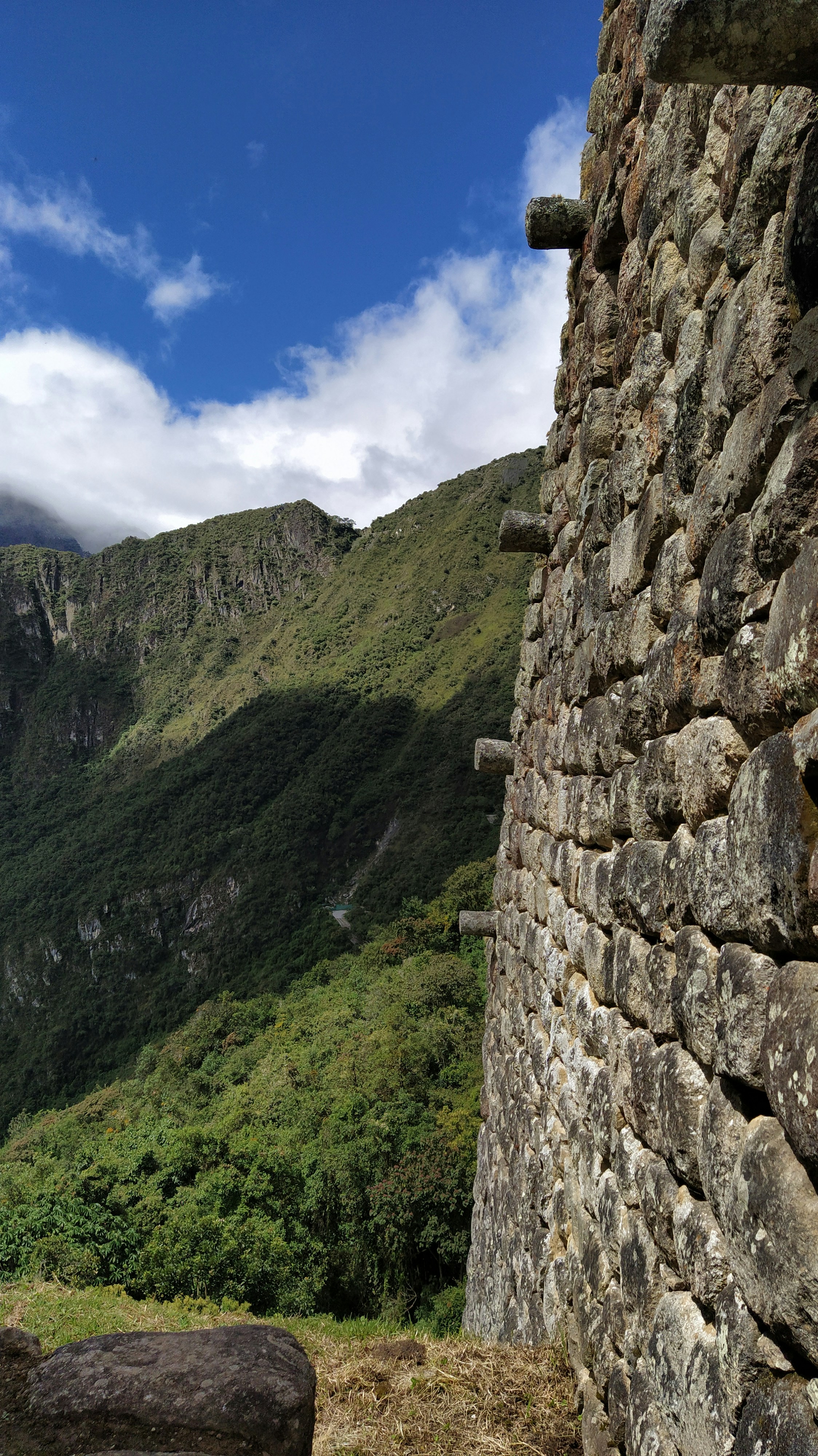 Rugged stone wall lines the right edge as a verdant valley and distant mountains unfold beneath a bright blue sky.
