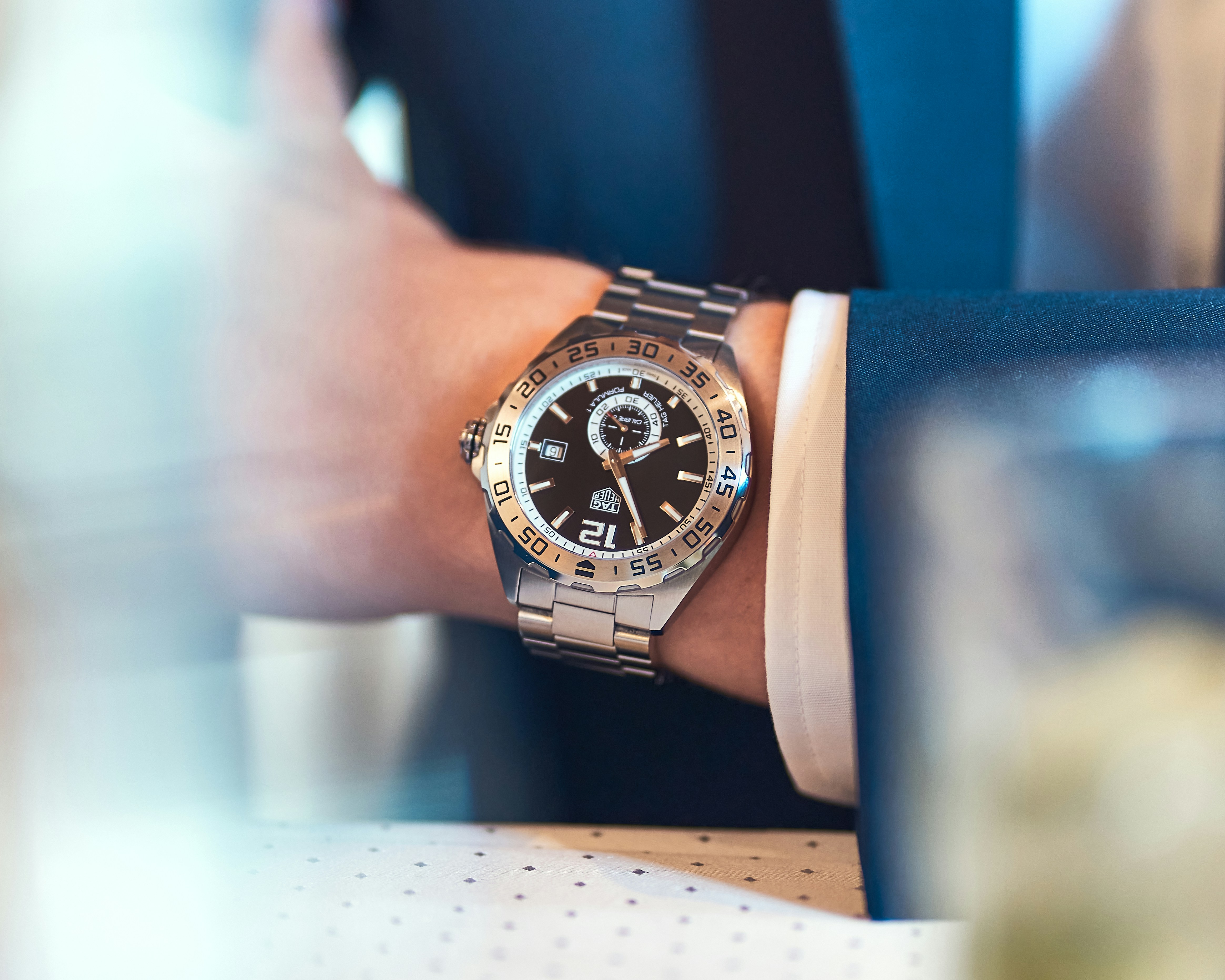Close-up of a sophisticated watch on a wrist, showcasing its intricate design and craftsmanship. The background features a blurred table setting, enhancing the focus on the timepiece.