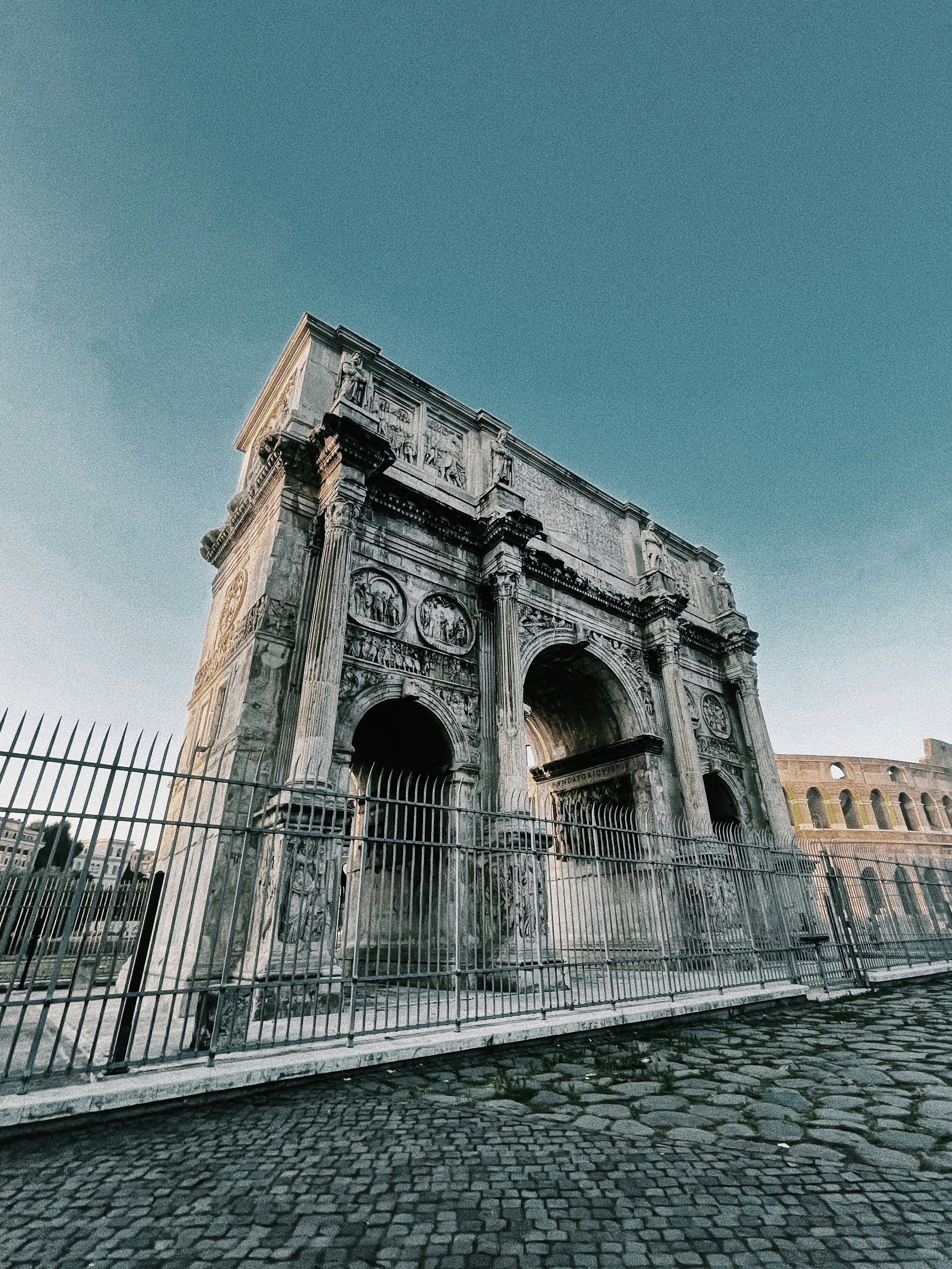 The Arch of Constantine stands majestically against a clear sky, framed by a wrought-iron fence and cobblestone pavement.
