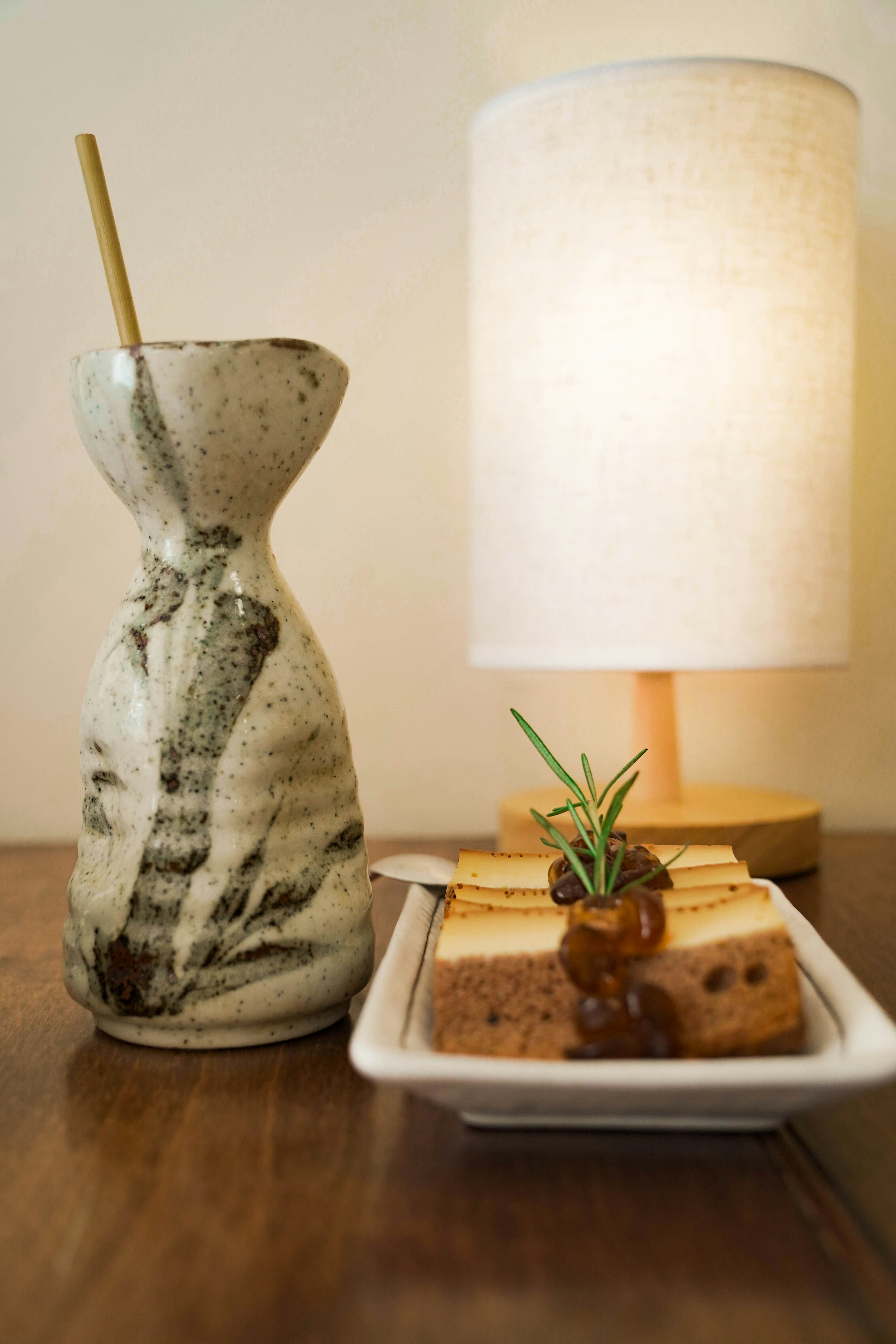 Ceramic marble-pattern carafe and a plate of square cakes with a rosemary sprig sit on a wooden table beside a warm lamp.
