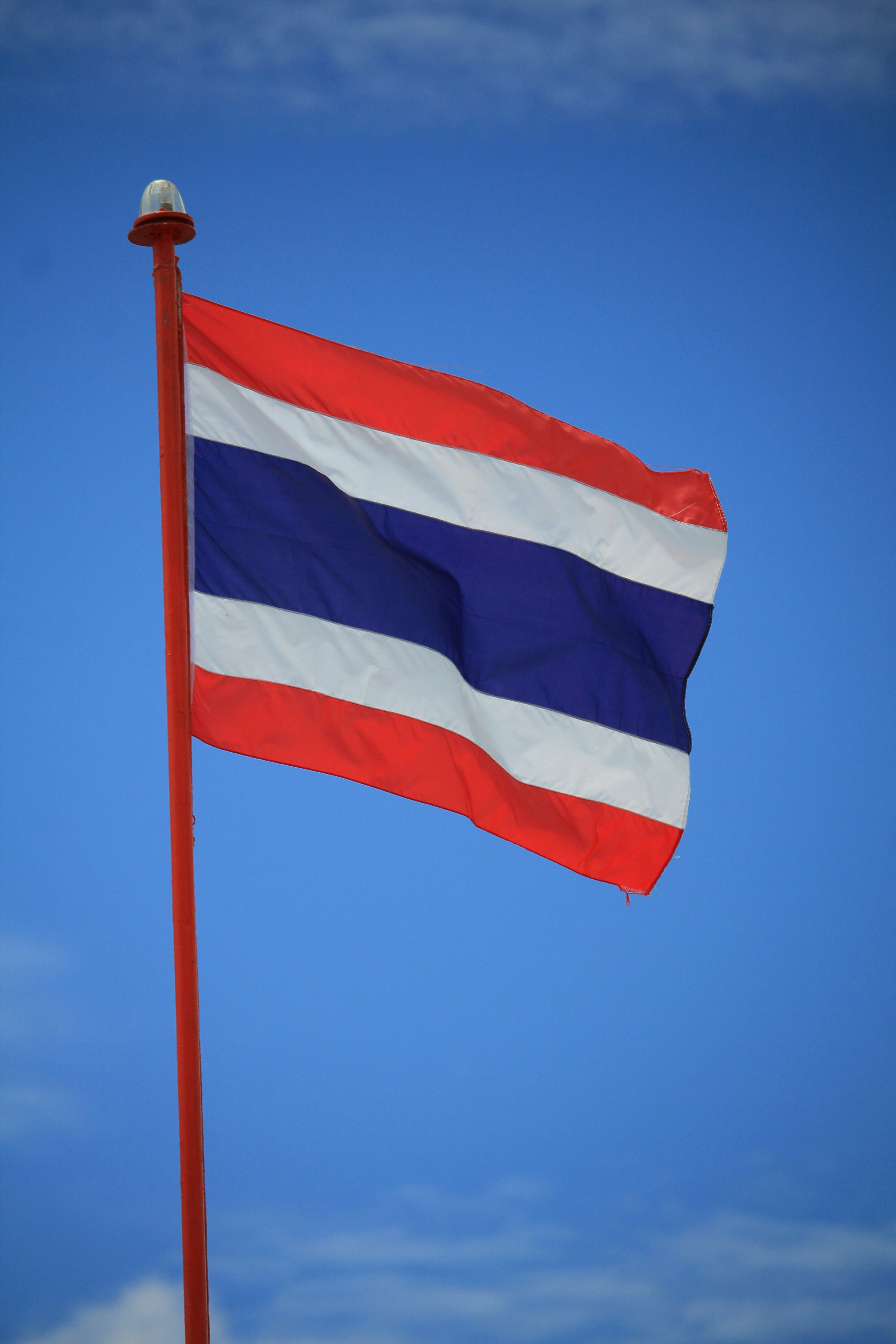 Thailand flag waving against a clear blue sky with scattered clouds.