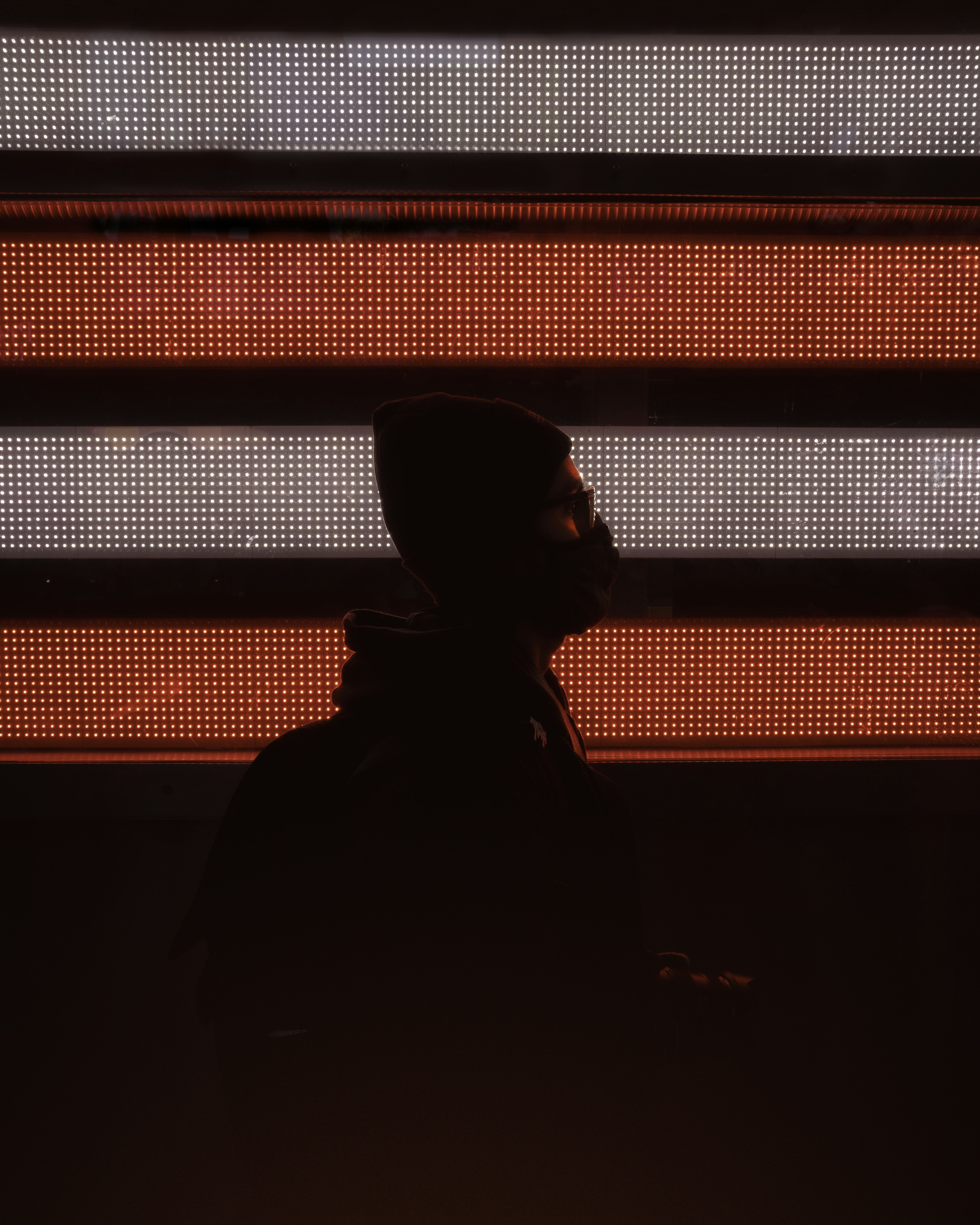 Silhouette of a person wearing a beanie and mask, set against vibrant horizontal neon light stripes in orange and white.