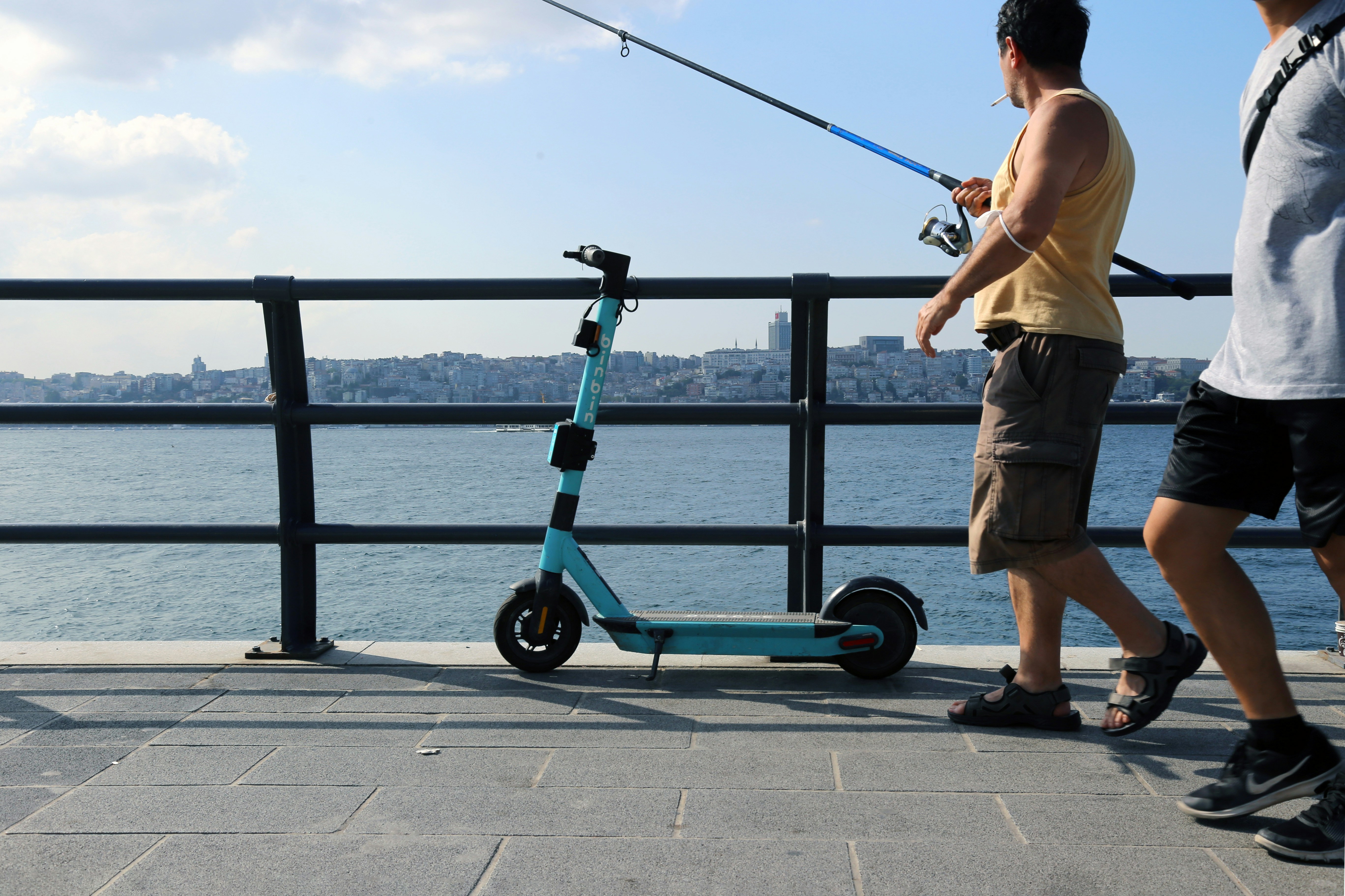 A man fishing from a waterfront promenade while a turquoise electric scooter stands nearby, illustrating the blend of urban lifestyle and leisure activities.