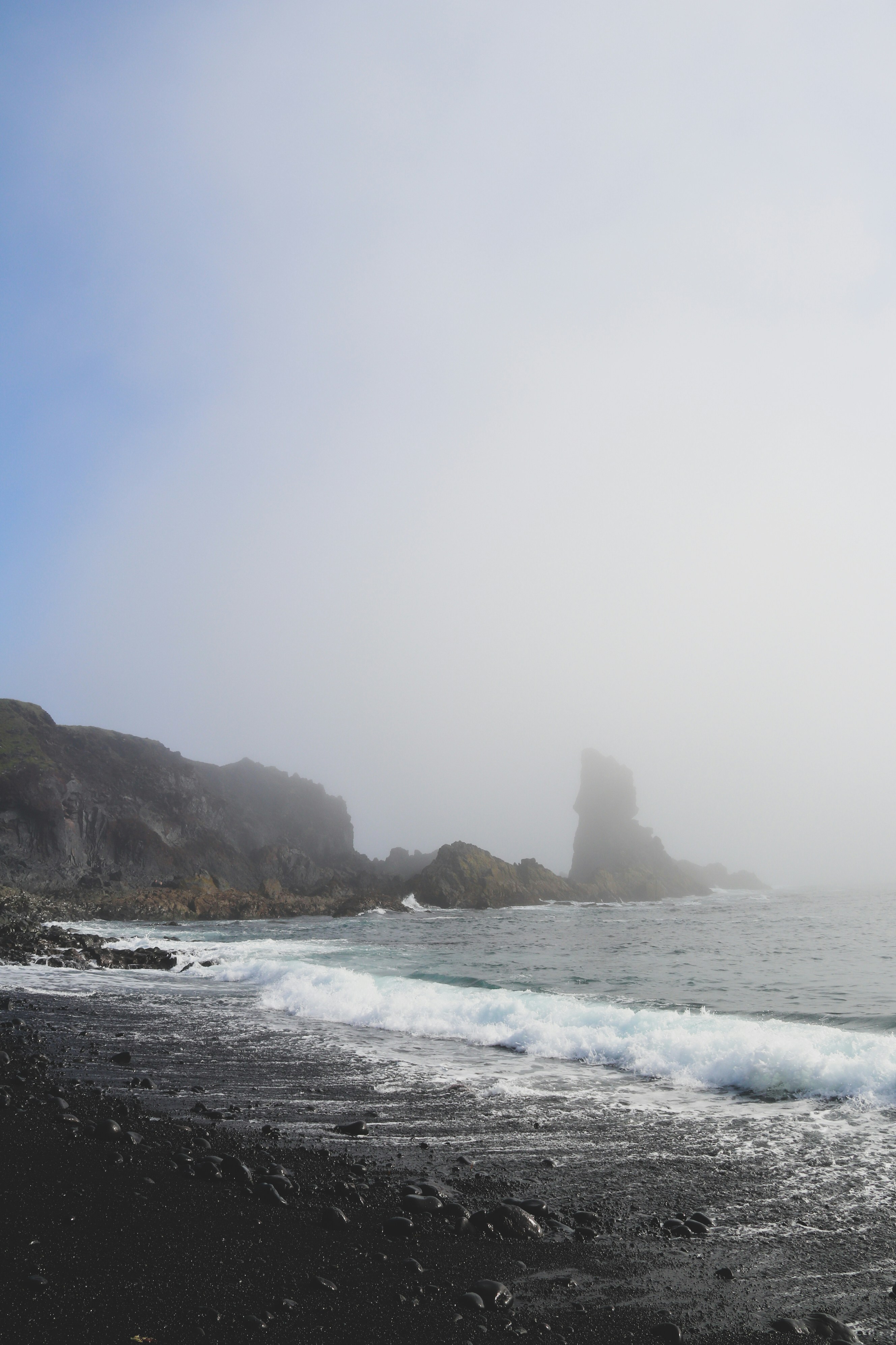 Fog envelops a rugged coastline, revealing a solitary rock formation amidst gentle waves on a black sand beach.