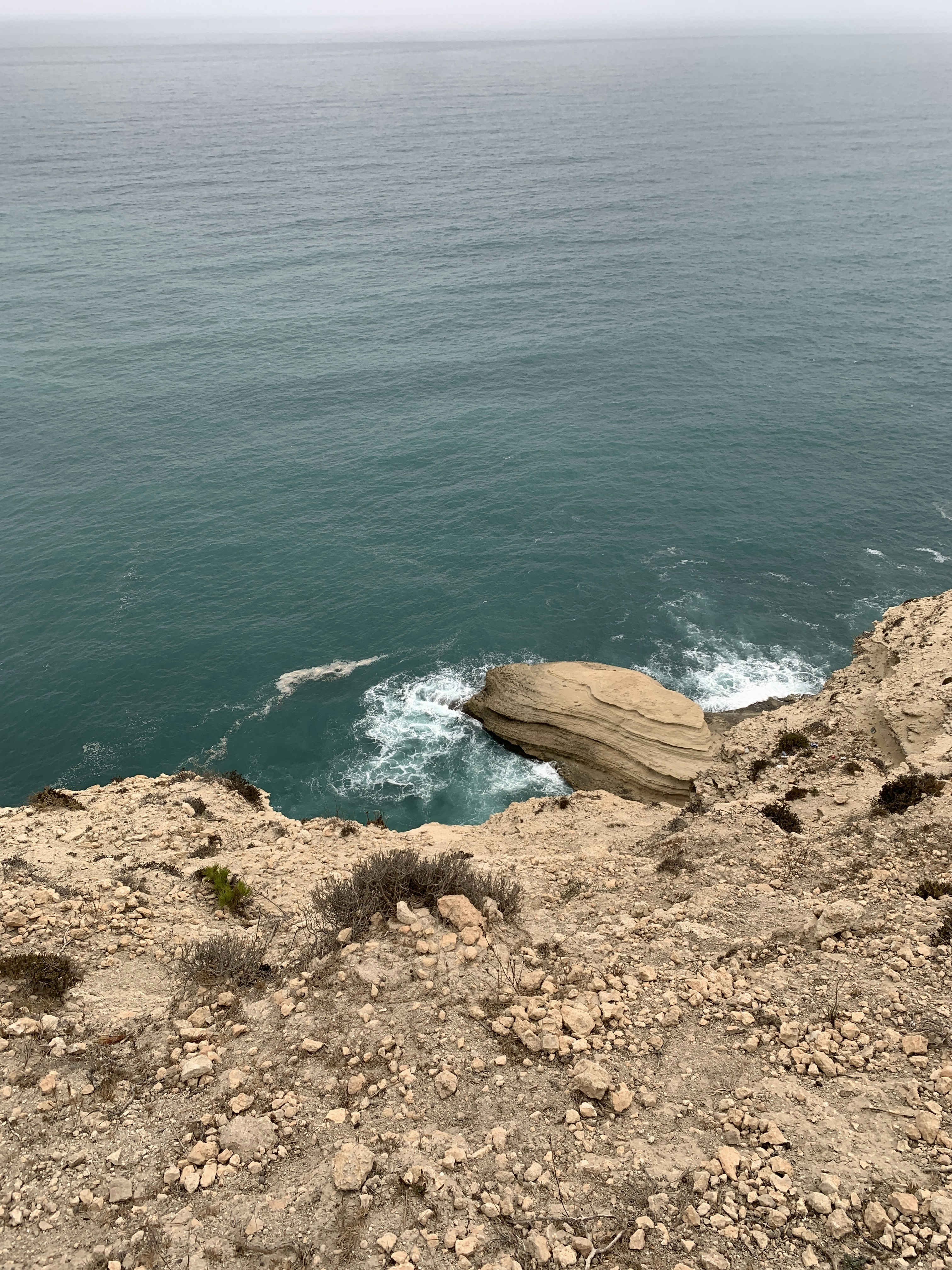Rocky cliff overlooking a vast, calm ocean with waves gently crashing on a lone rock.