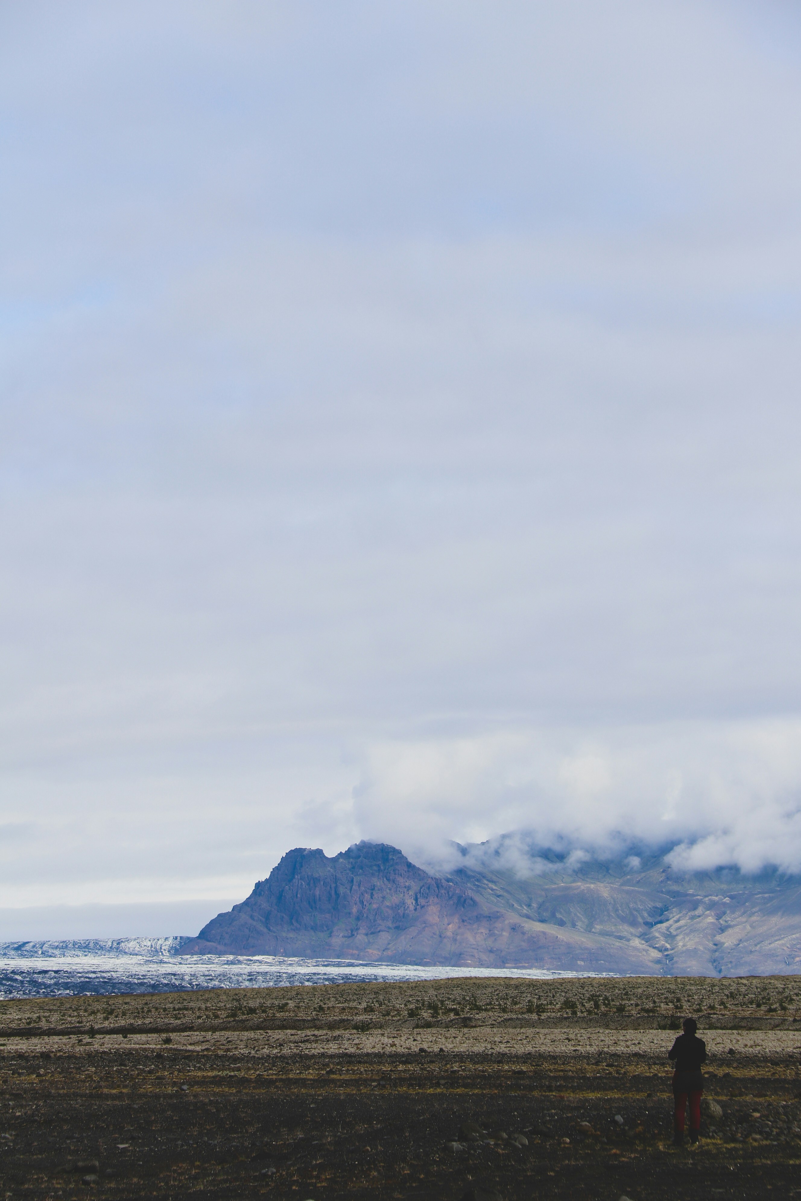 Red-clad silhouette at the edge of a wind-swept shoreline beneath cloud-blanketed peaks