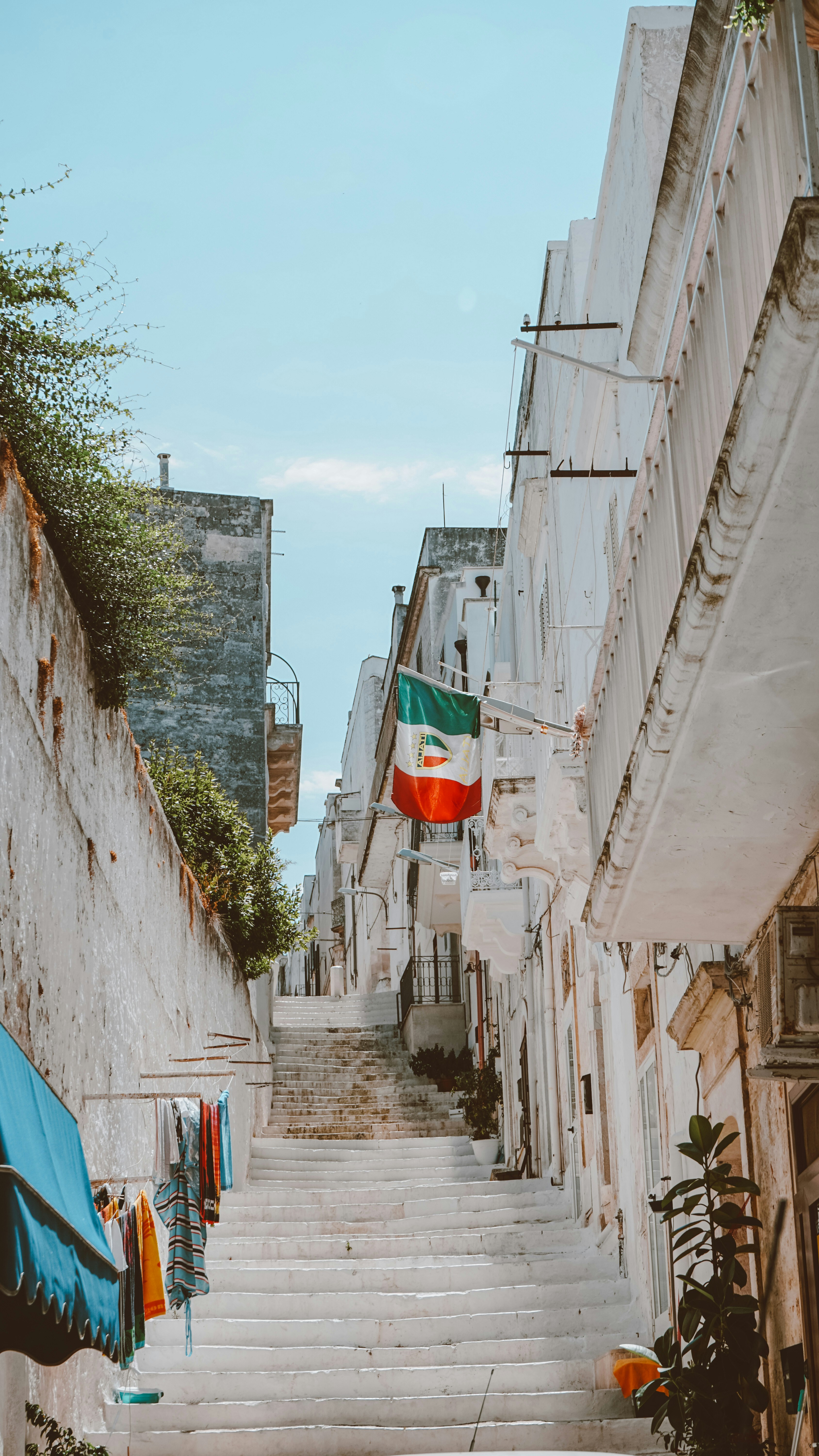 Colorful laundry hangs alongside a staircase leading up to a vibrant Mexican flag, framed by whitewashed buildings under a clear blue sky.