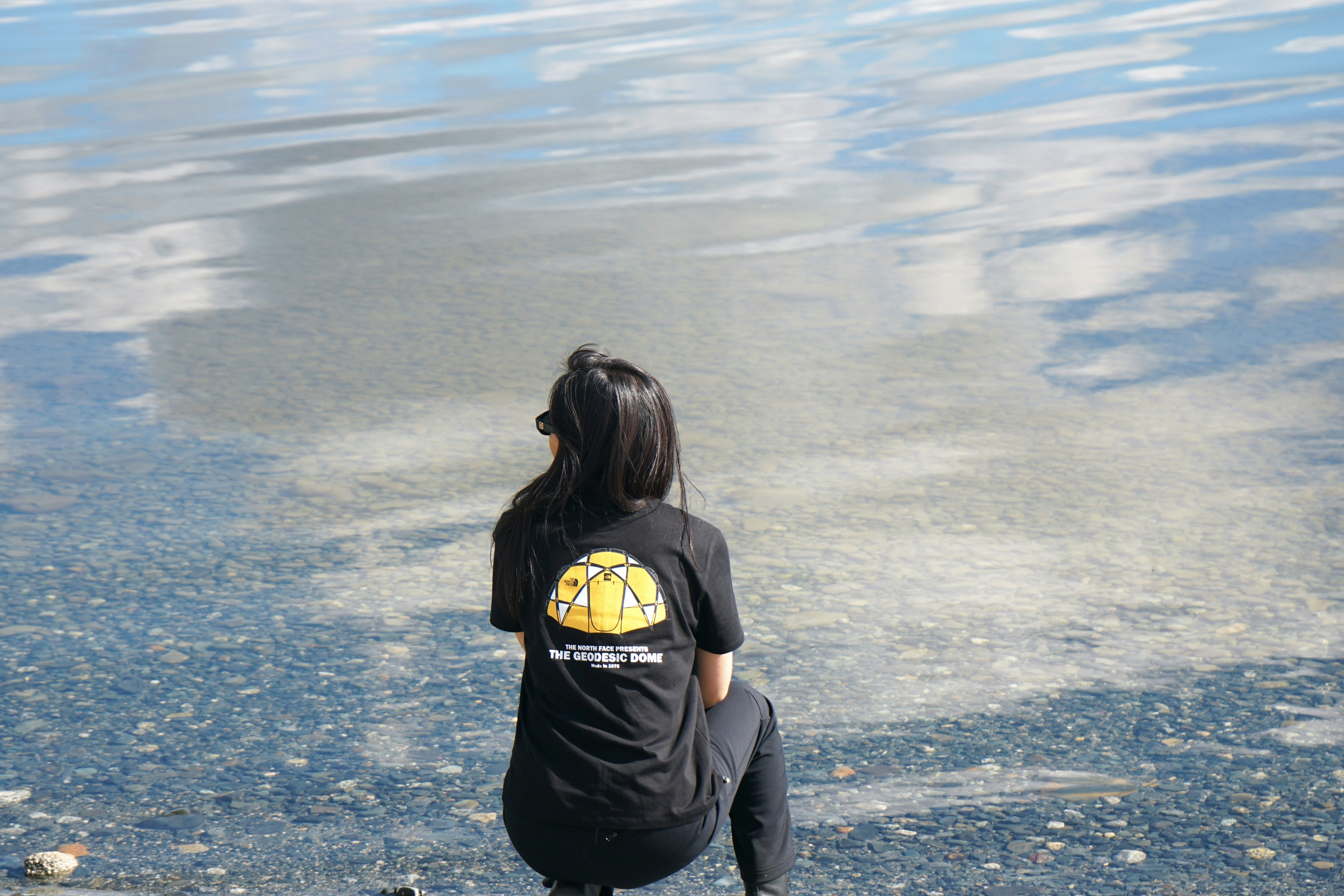 Person seated by the water, gazing at the reflective surface, wearing a black t-shirt with a yellow design. The serene environment captures a moment of contemplation.